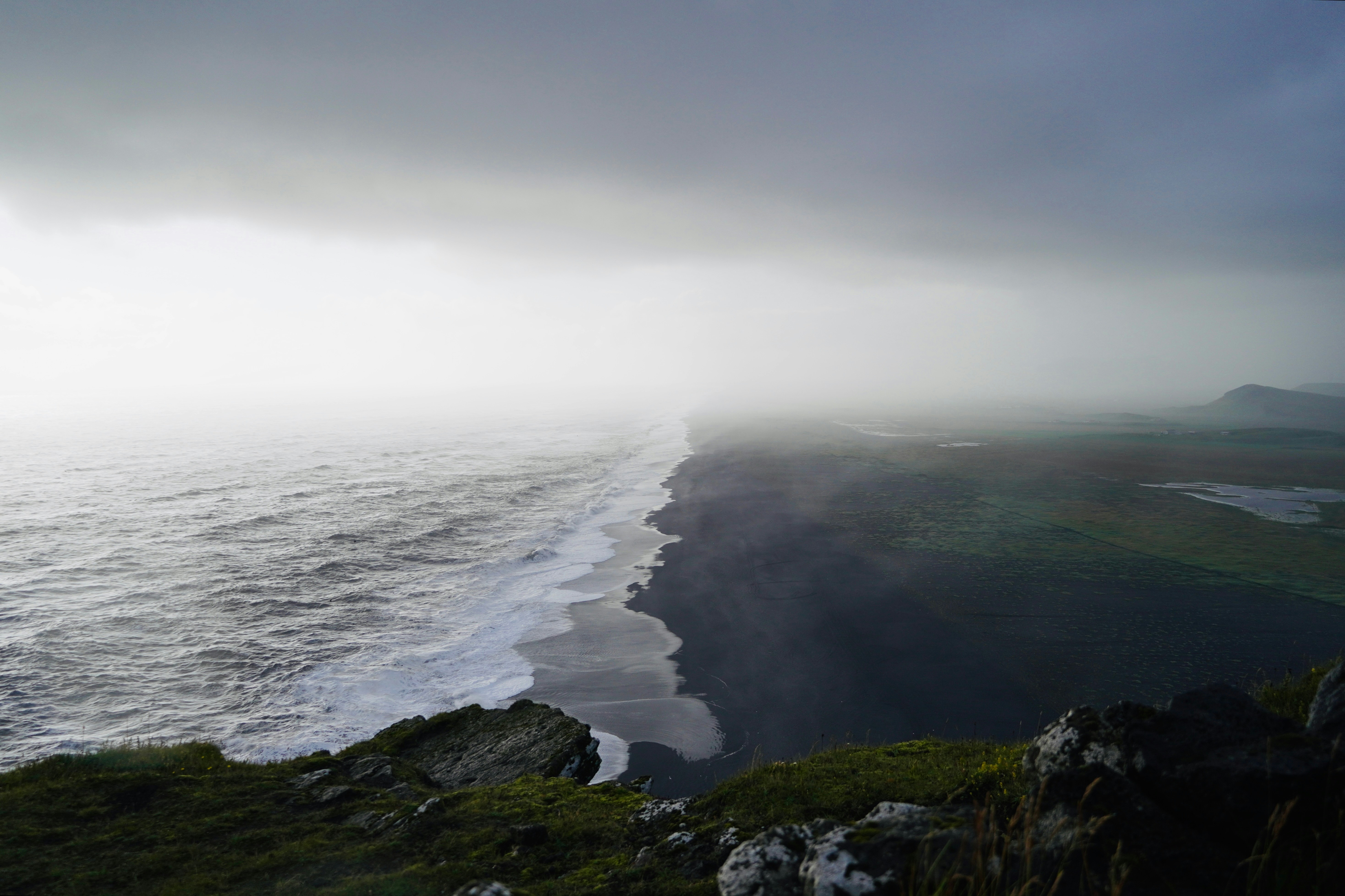Misty coastline with waves gently lapping against a dark shore, framed by lush green land under a dramatic sky.