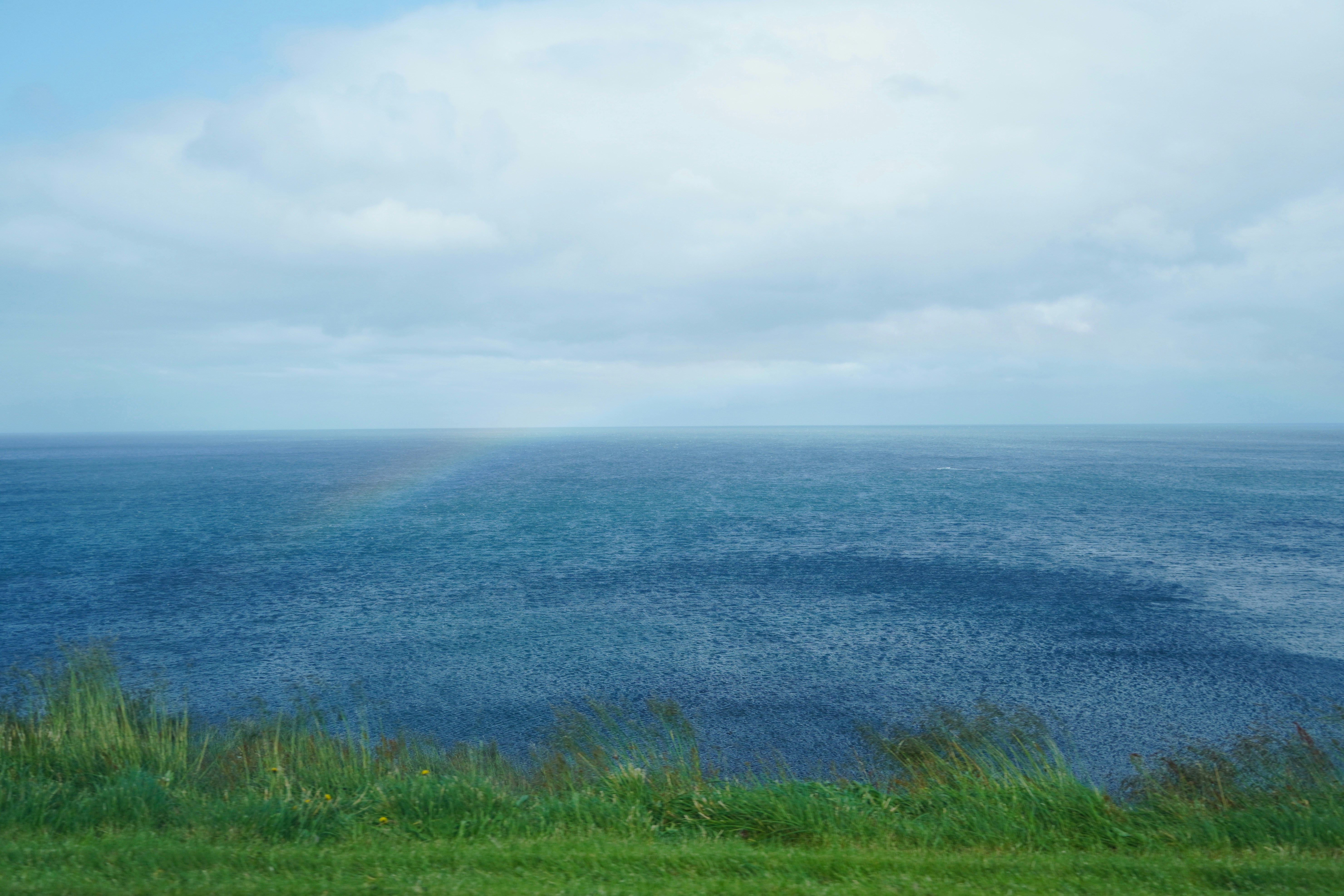 Gentle waves ripple across a vast ocean under a cloudy sky, with a faint rainbow emerging in the distance. Lush grass frames the foreground.