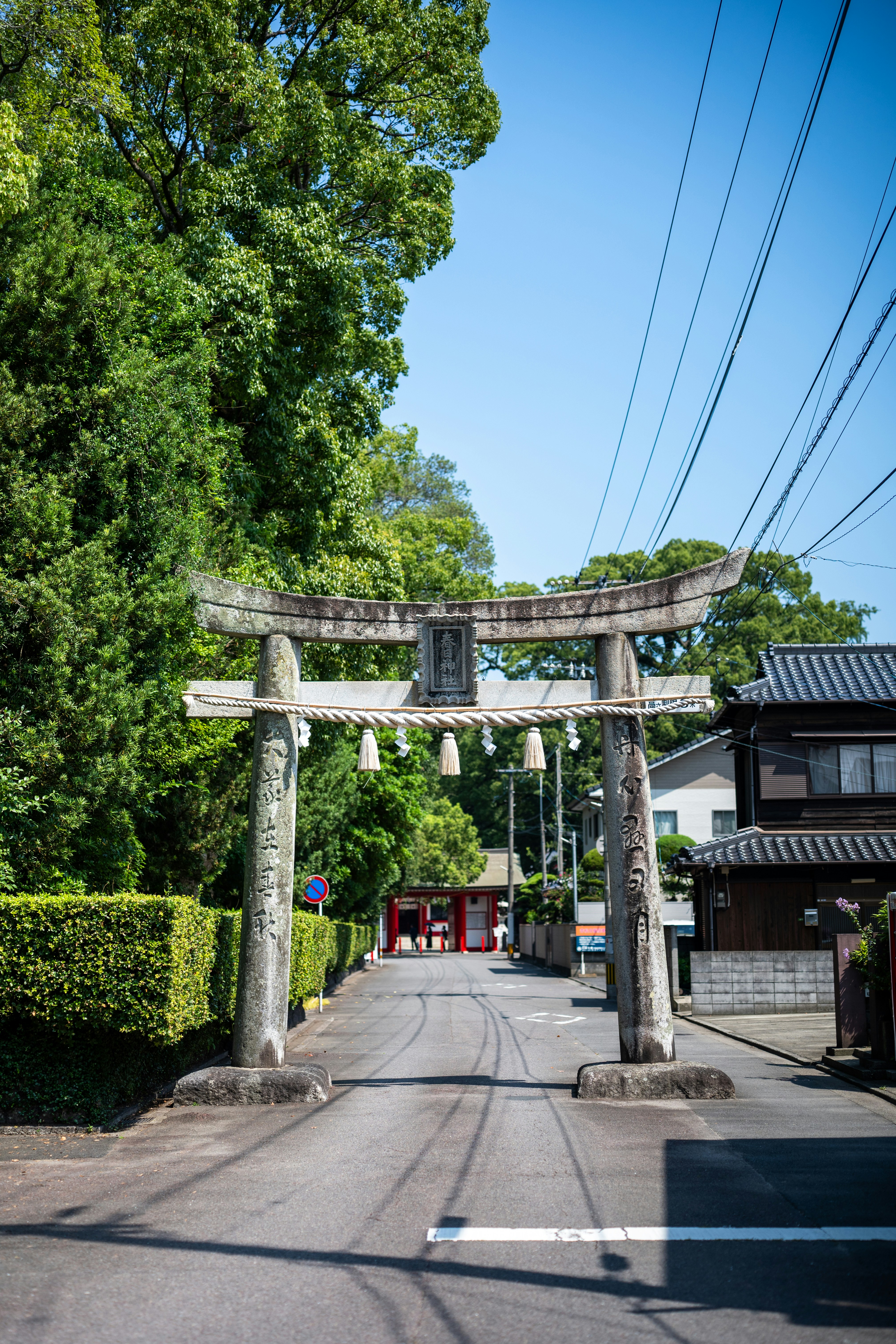 Traditional torii gate marking the entrance to a serene path lined with lush greenery and residential buildings. The clear blue sky enhances the peaceful atmosphere.