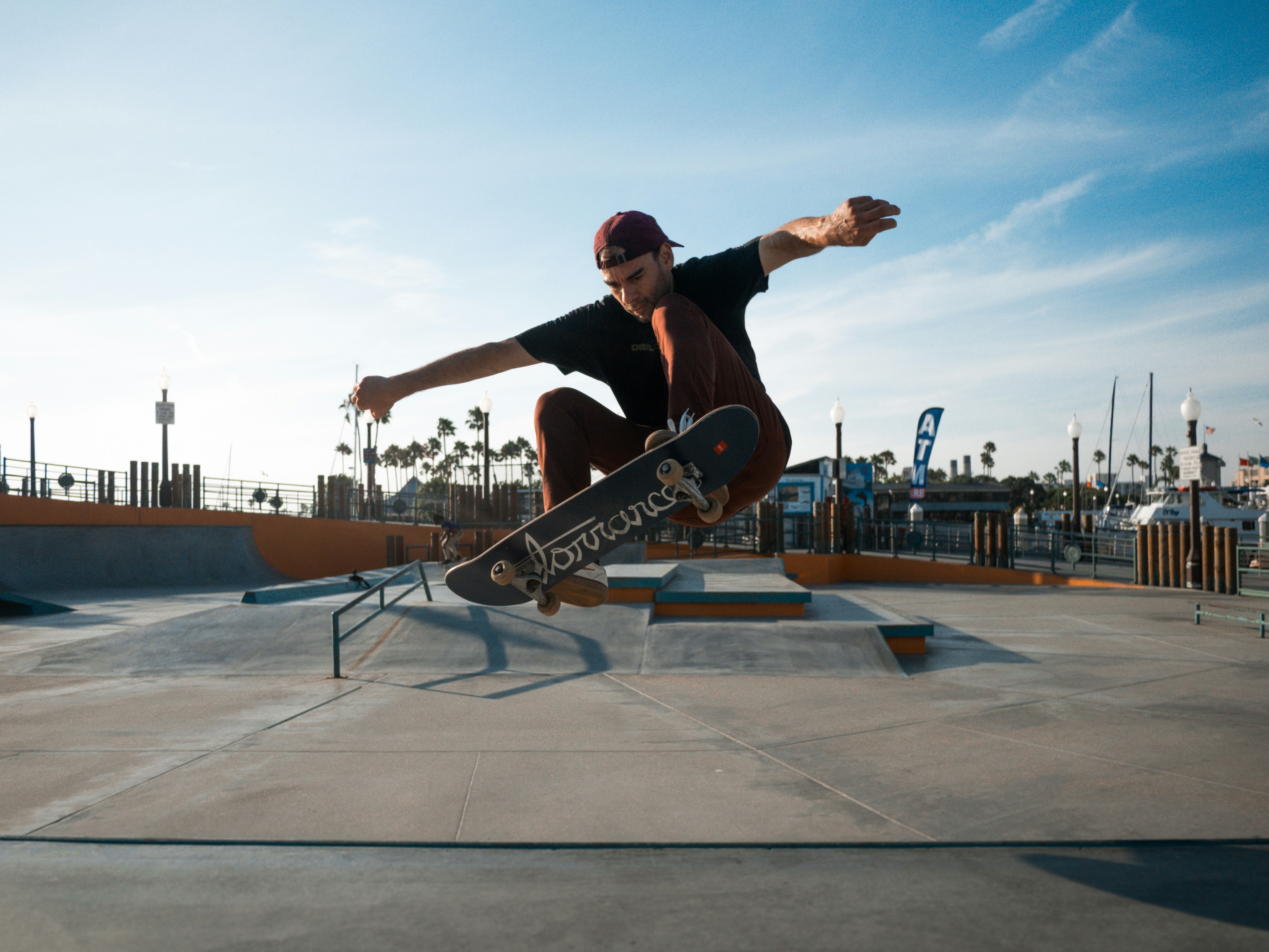 Skateboarder executing a mid-air trick above a concrete skate park with palm trees in the background.