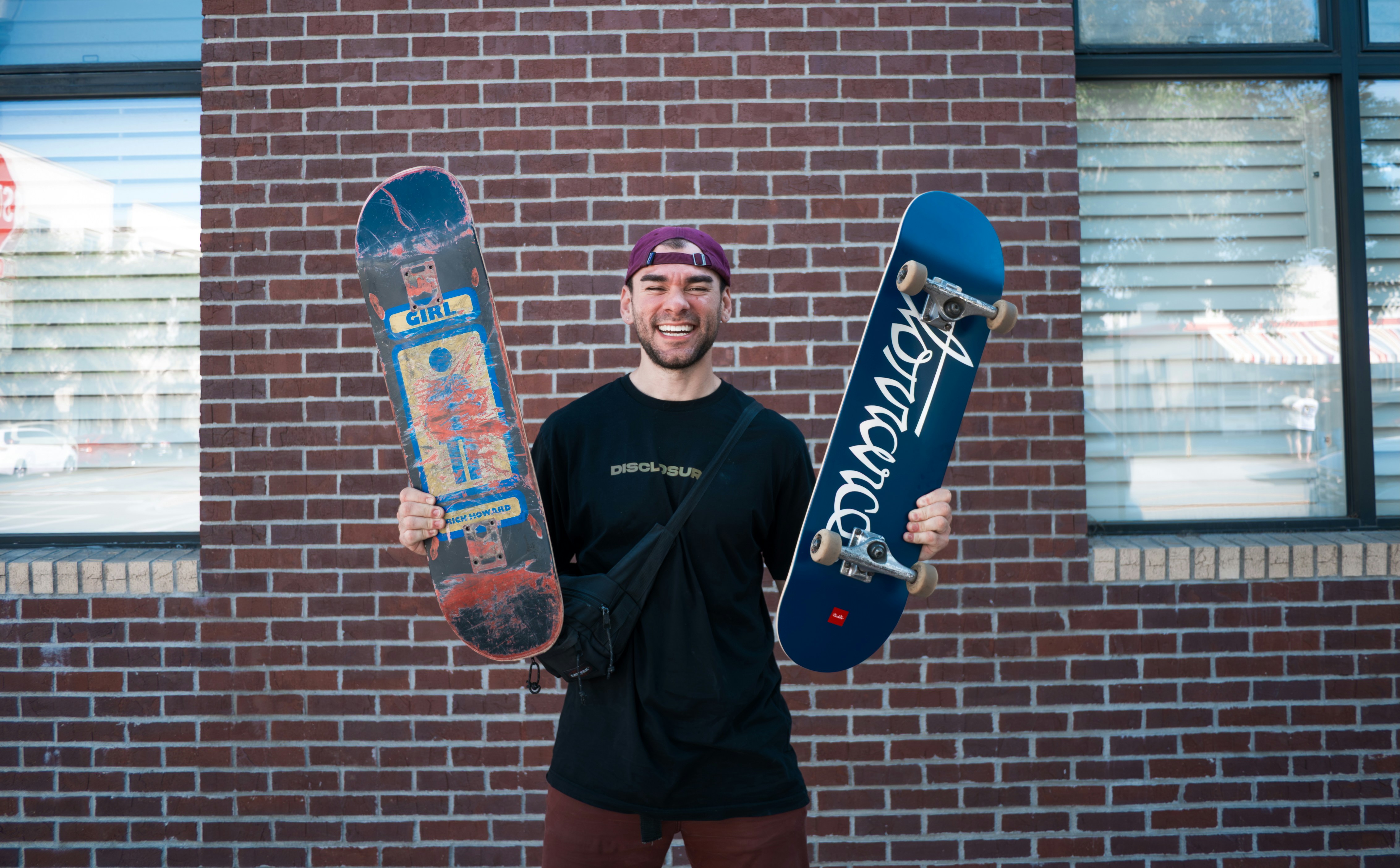 Enthusiastic skater proudly displays two skateboards, showcasing his love for the sport against a brick wall backdrop.