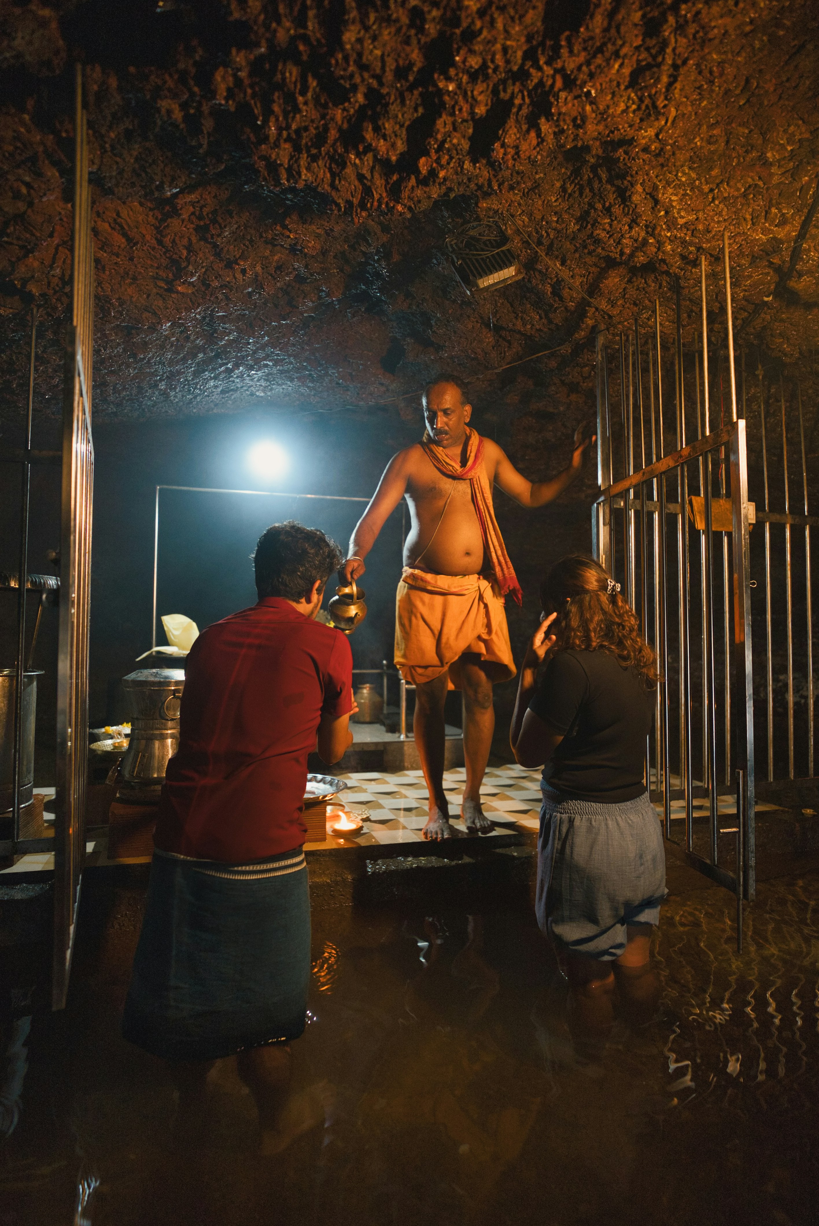 A priest performs a ritual in a dimly lit cave, surrounded by water, while two devotees observe with reverence. The atmosphere is steeped in spirituality.