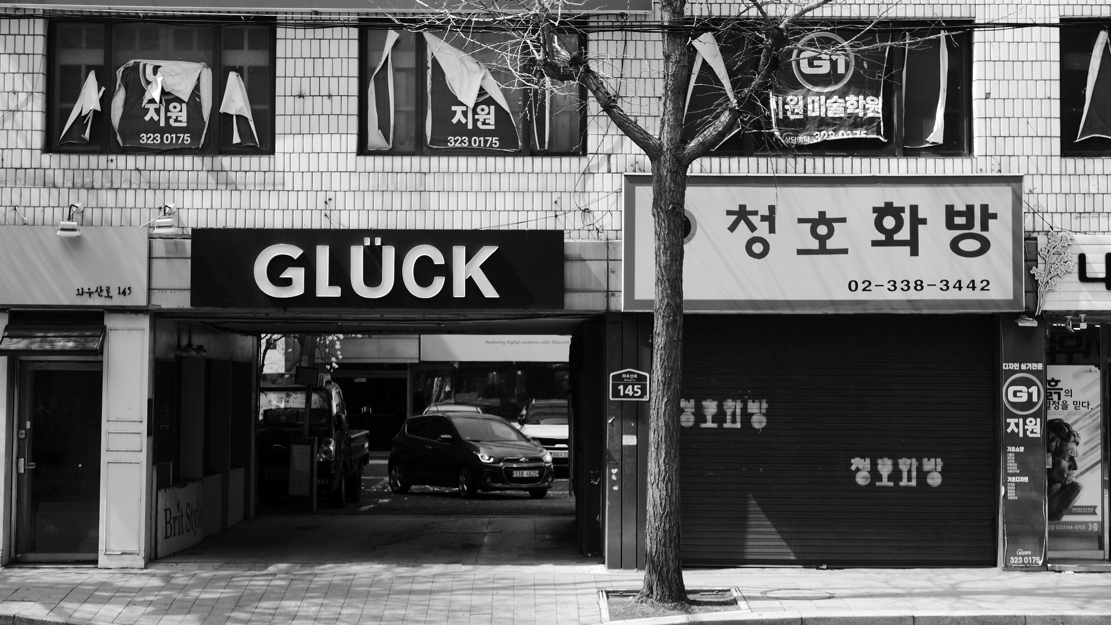 A monochrome street scene featuring the sign 'GLÜCK' prominently displayed, surrounded by various storefronts and a tree, evoking a sense of calm in the urban environment.