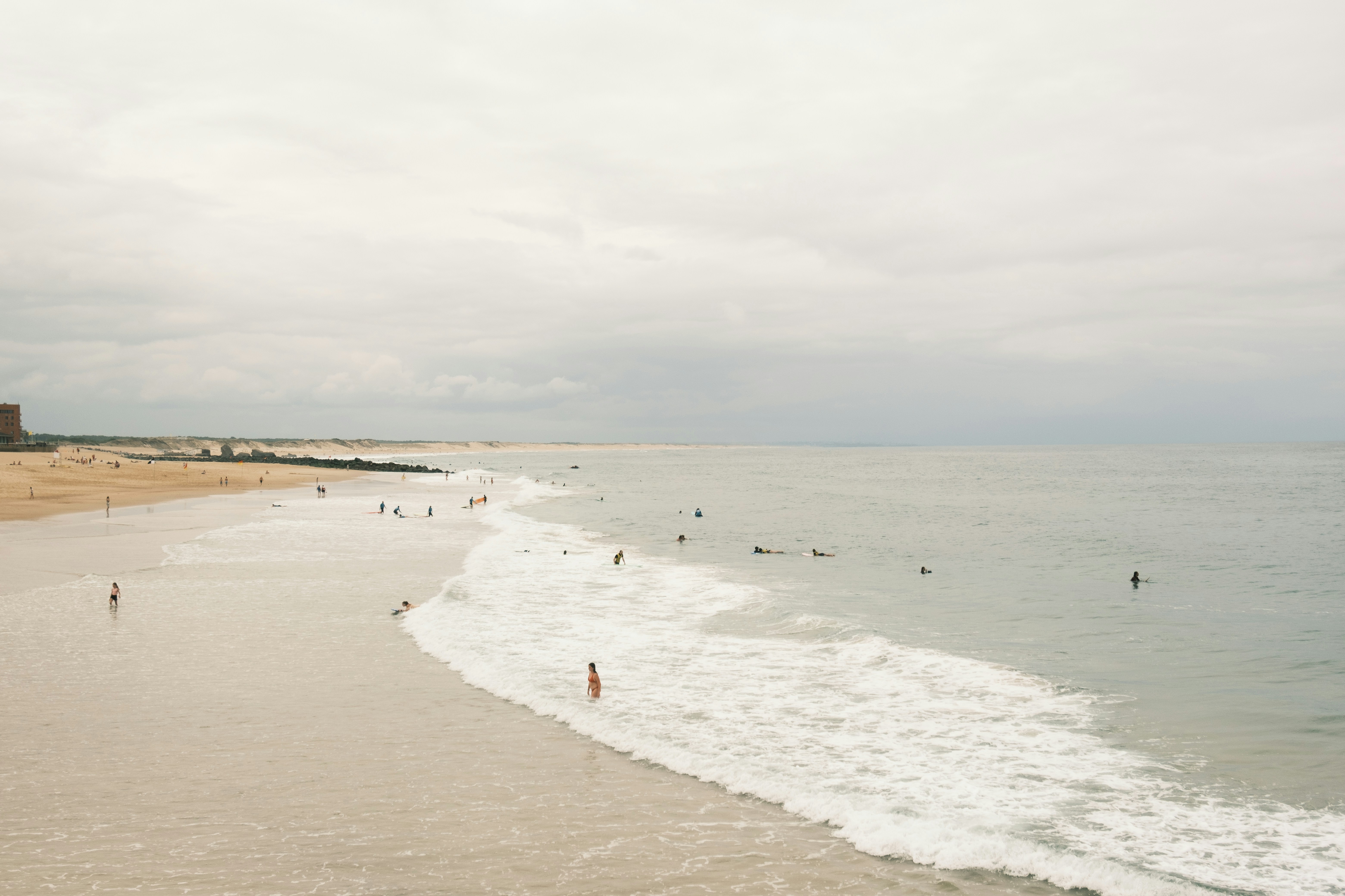 Surfers and beachgoers enjoy a tranquil day at the shoreline, where gentle waves lap against the sandy beach under a cloudy sky.