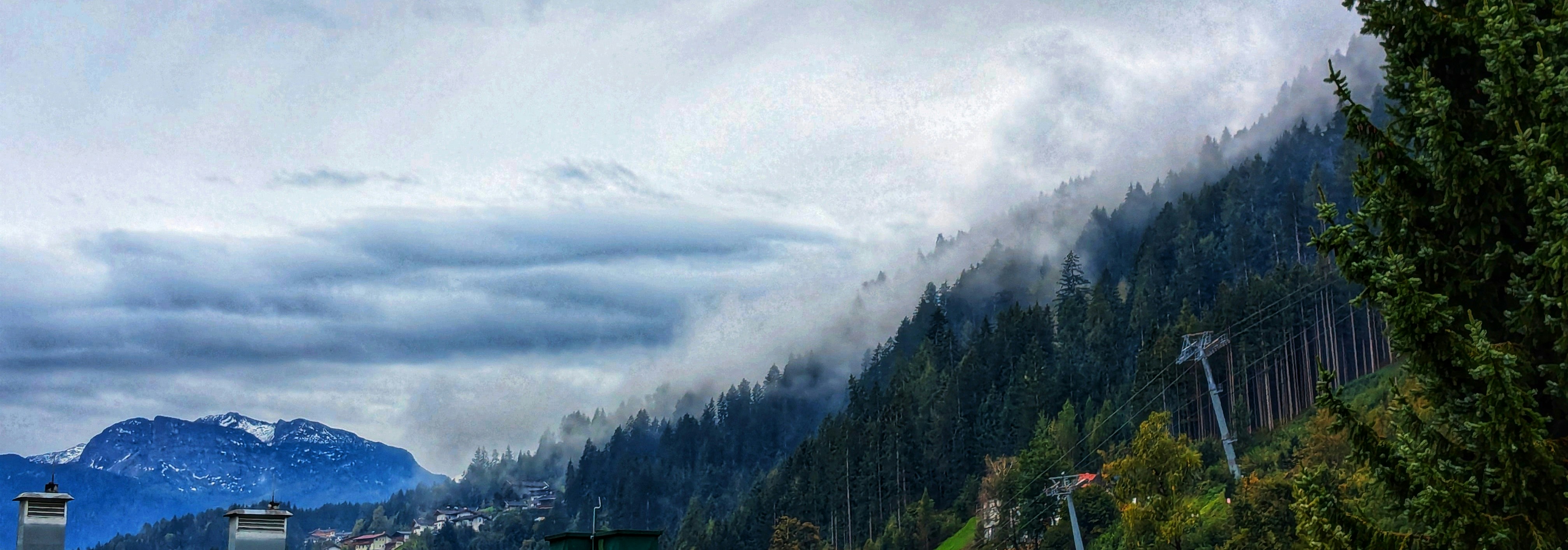 A dense fog envelops the mountainside, blending with the lush greenery and hinting at the beauty beyond. The scene captures the essence of a tranquil alpine landscape.