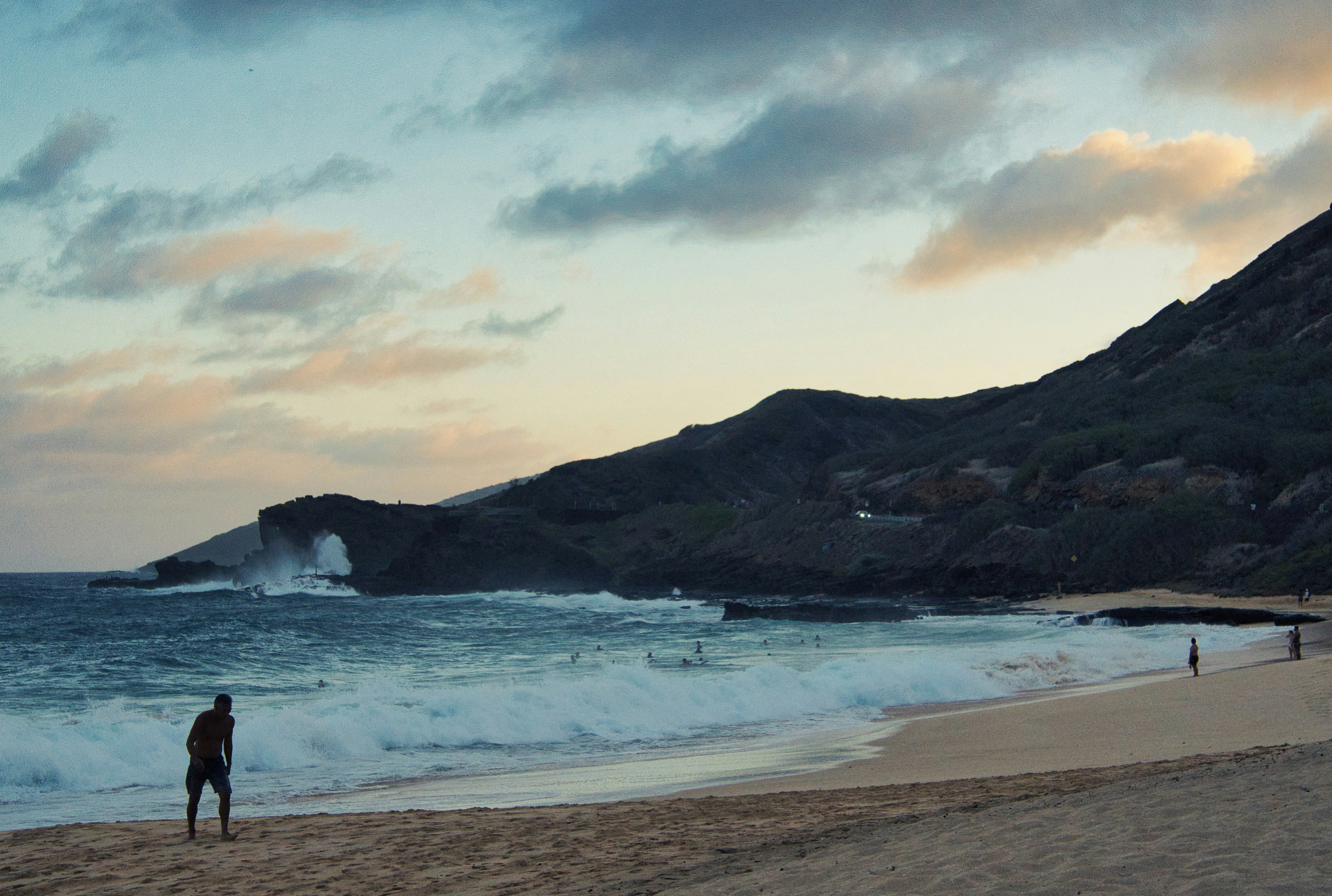 A solitary figure walks along a sandy beach as gentle waves lap at the shore, with a rugged coastline and soft clouds in the backdrop.