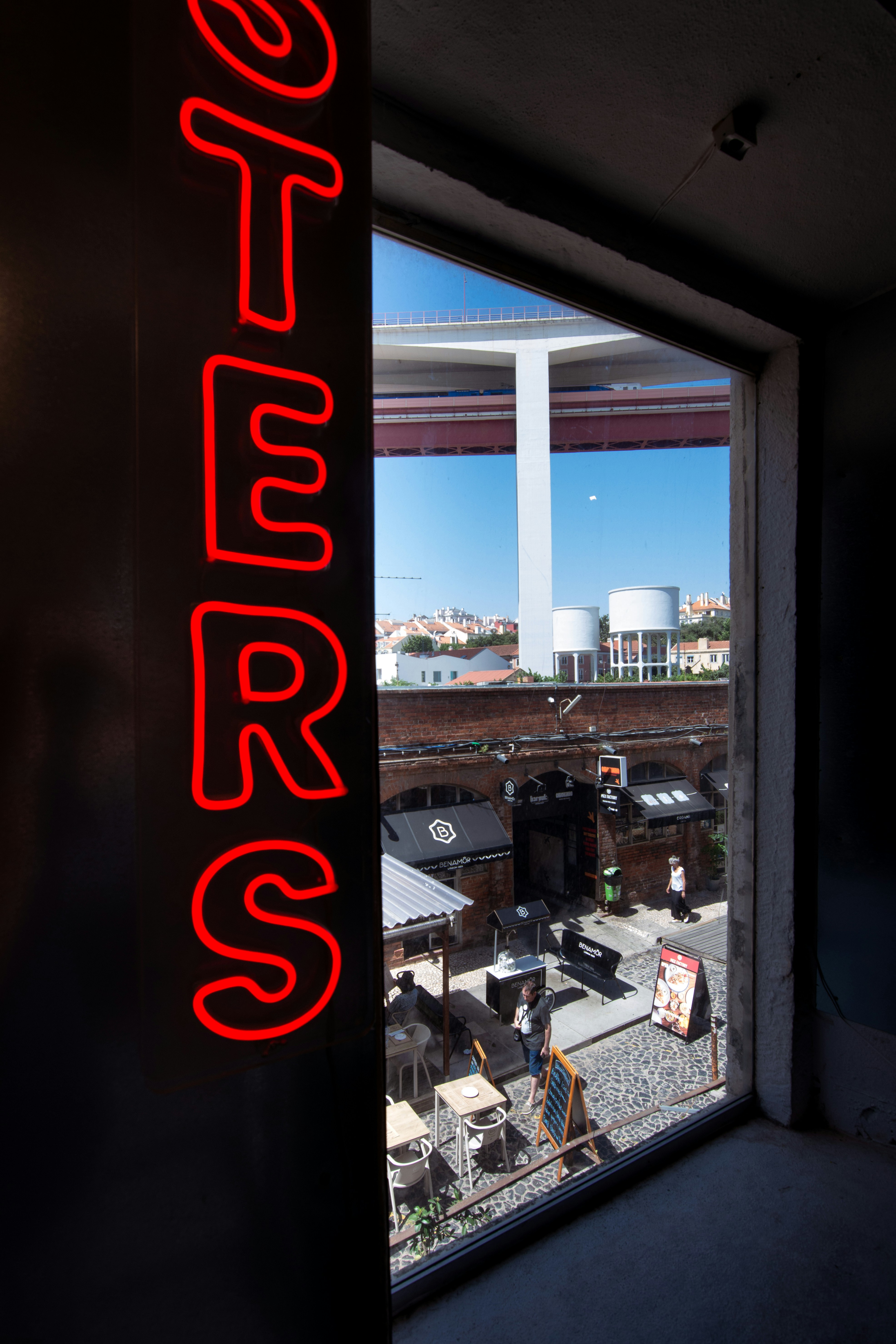 Neon sign for 'Oysters' framed by a window, overlooking a bustling outdoor market with tables and a bridge in the background.