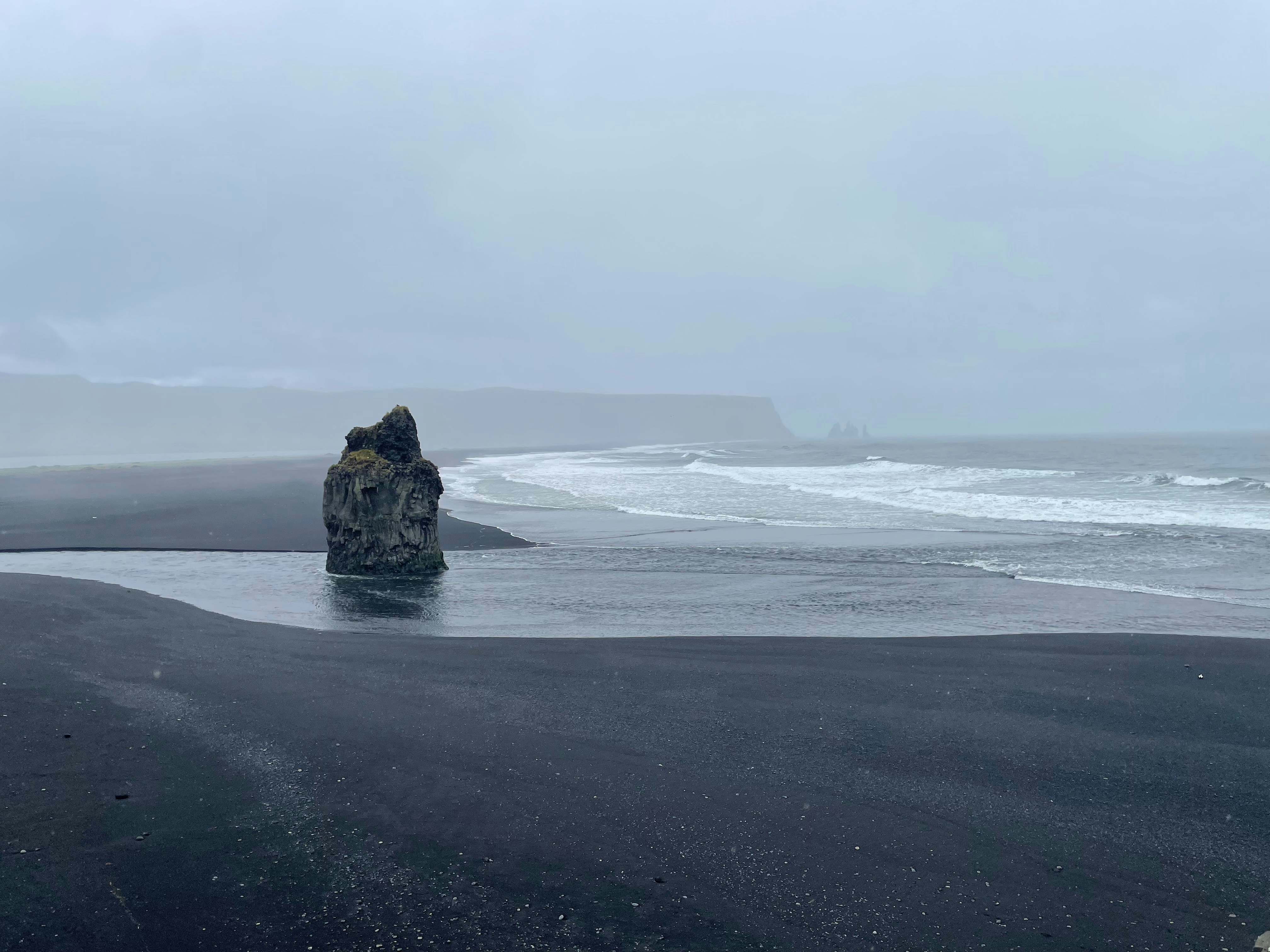 Large rock formation on a black sand beach with waves.