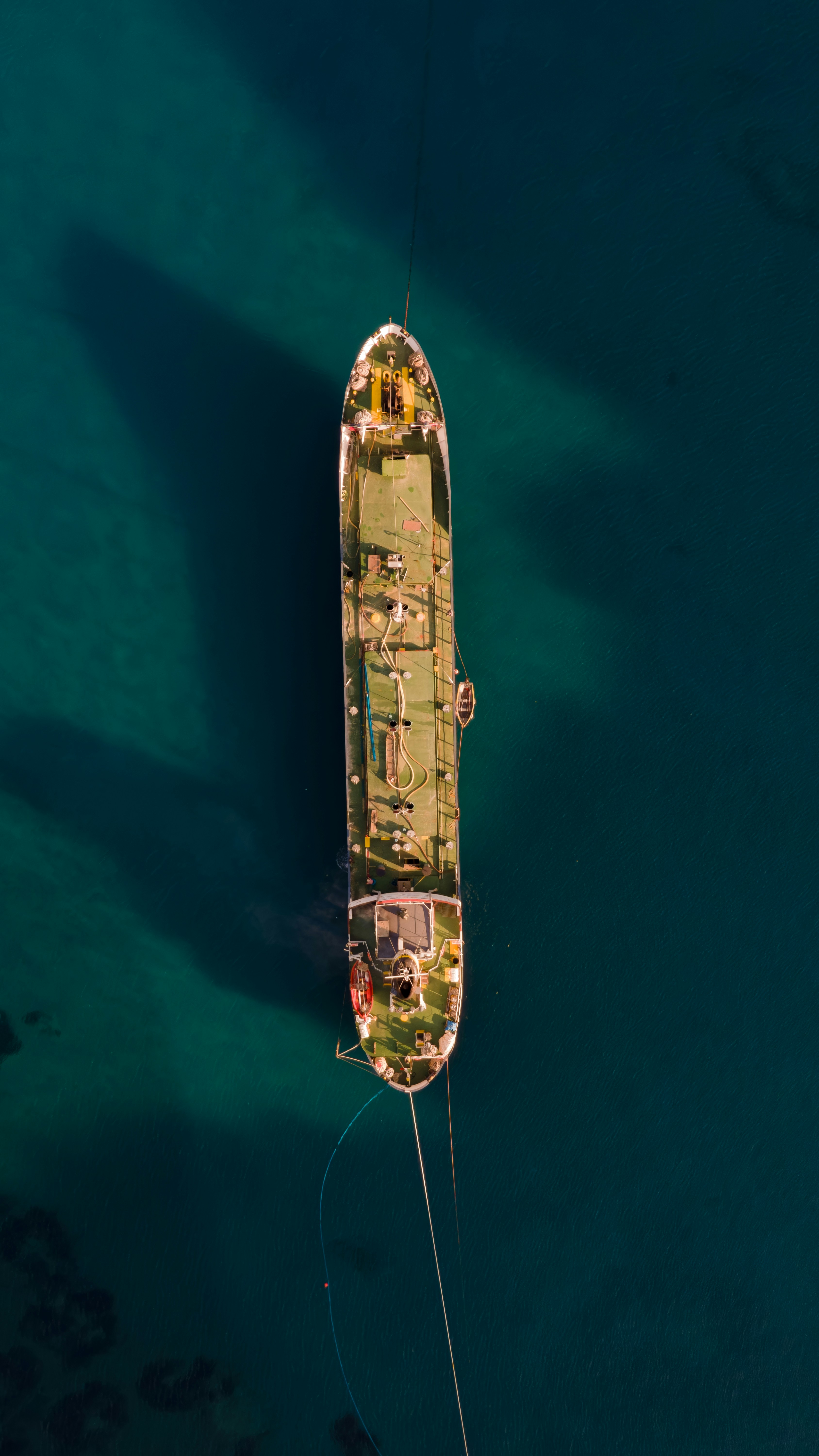 Aerial perspective of a cargo ship anchored in tranquil waters, showcasing its intricate deck layout and mooring lines. The vibrant blue sea contrasts with the ship's green surface.