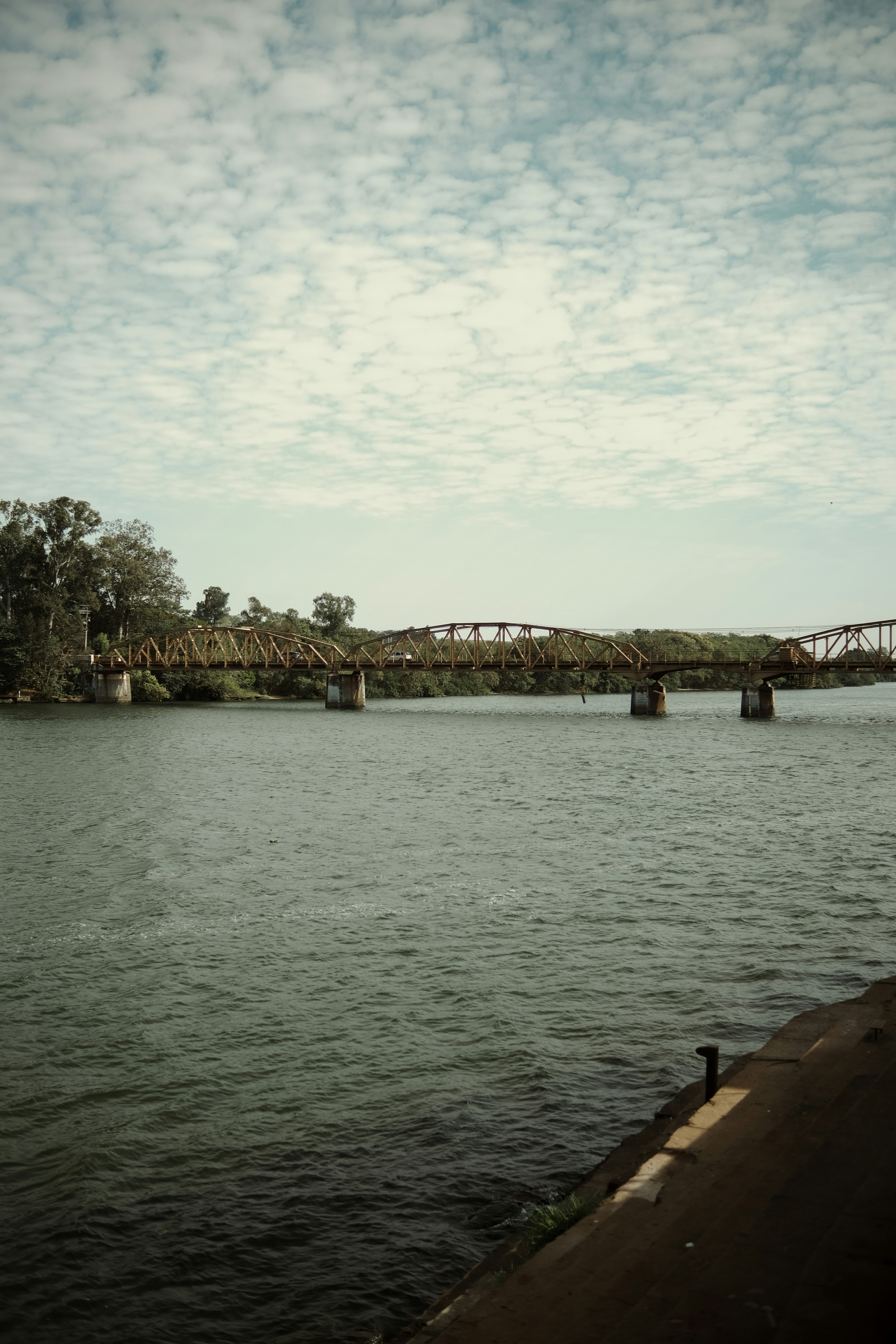 Old metal bridge crossing a wide river under cloudy sky