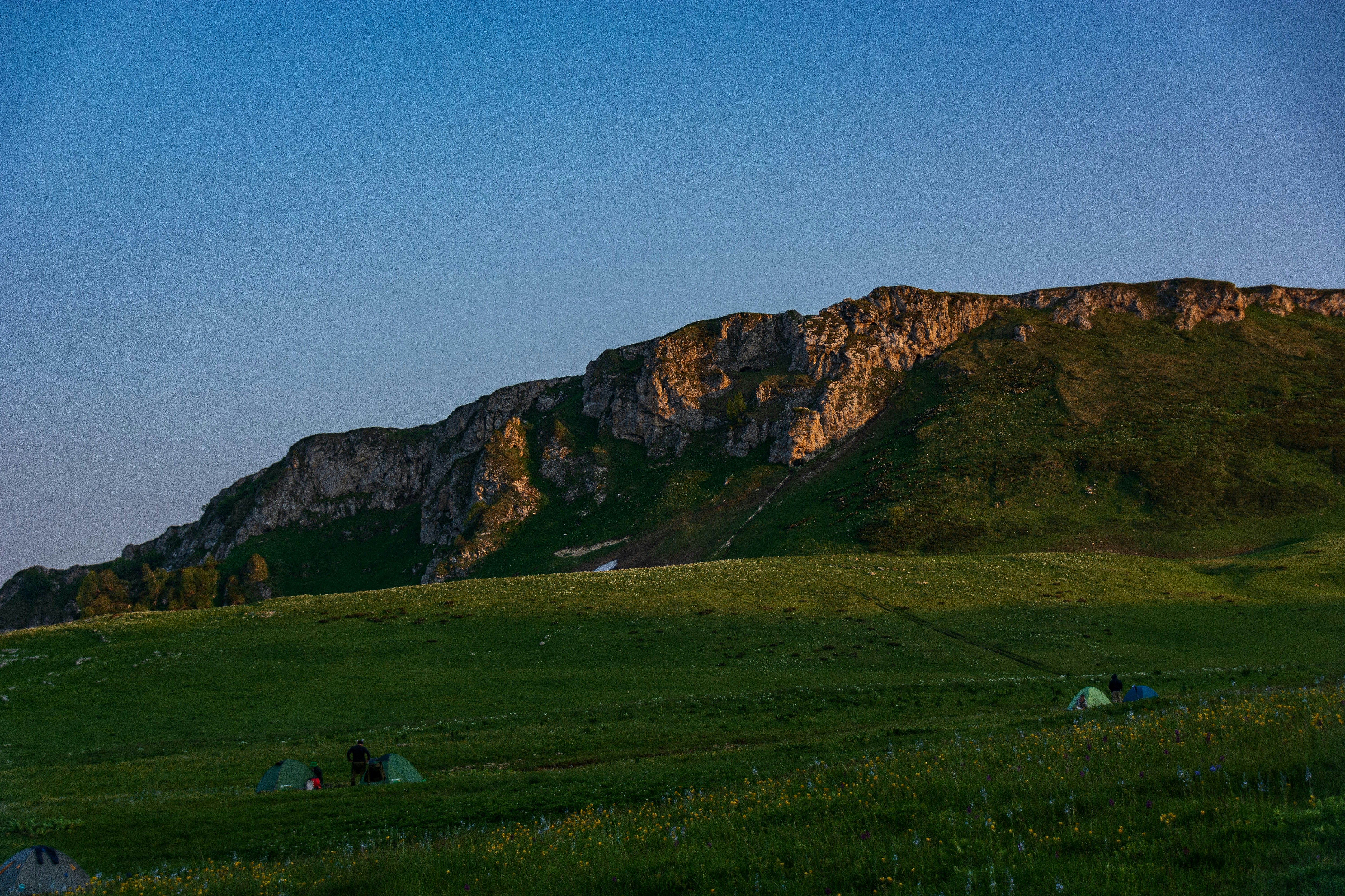 Green grassy hills with tents and rocky mountain at sunset