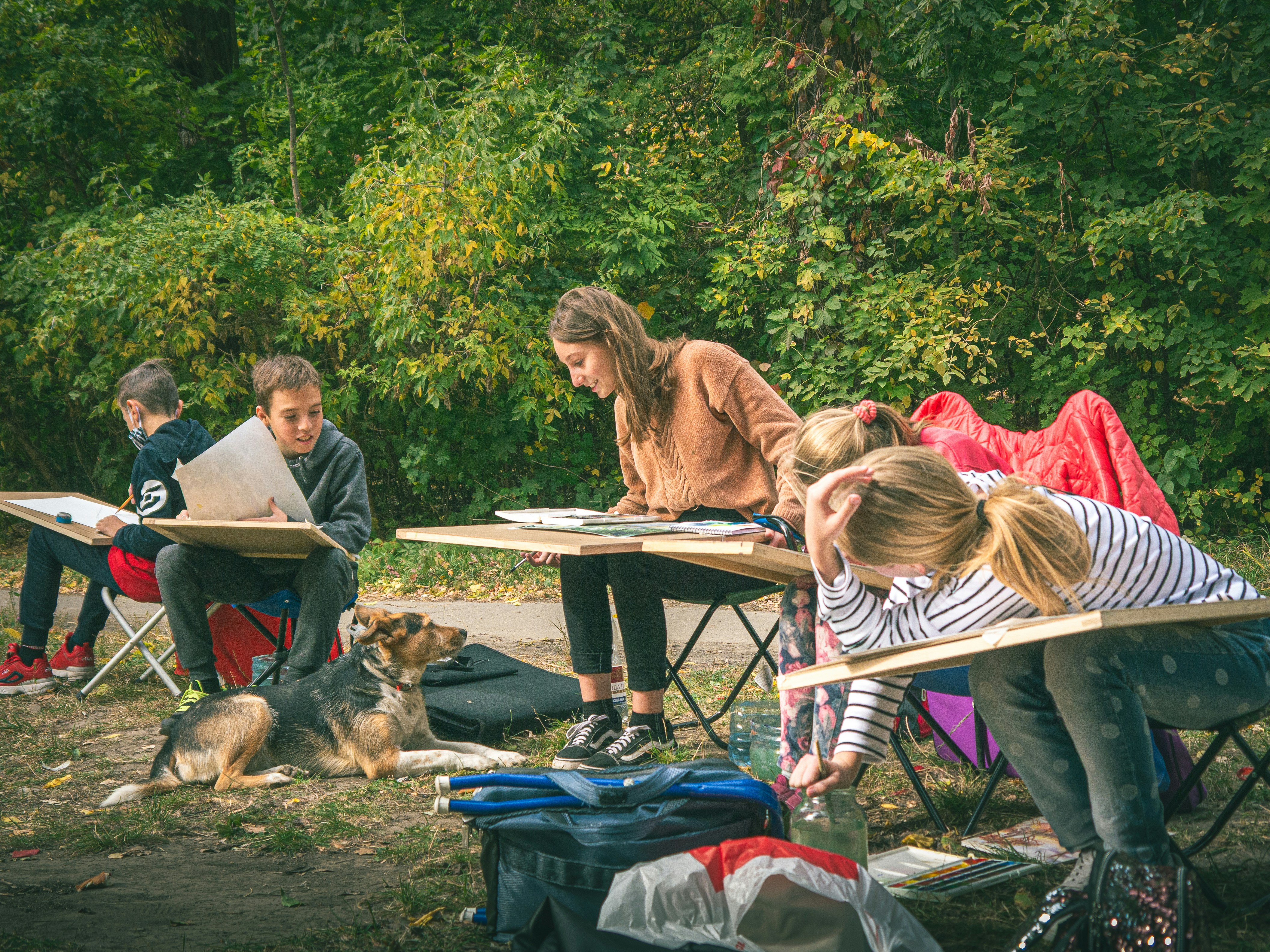 A candid moment of outdoor creativity — children absorbed in drawing, supervised by an unexpected but very loyal companion. Shot in natural light with a documentary feel. | People sketching outdoors with a dog nearby