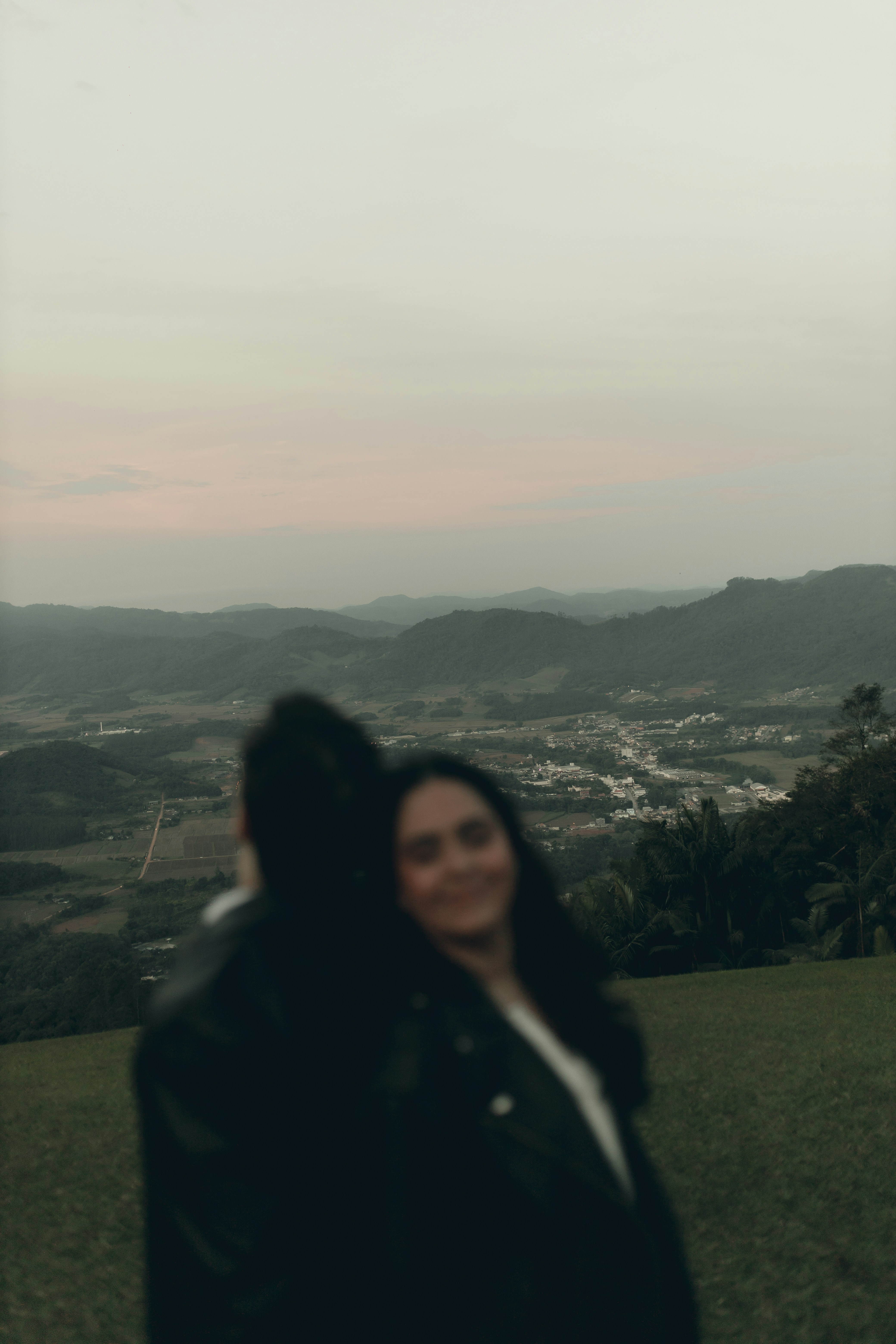 Two figures in leather jackets stand in the foreground, partially blurred, against a sprawling landscape at dusk, with mountains and a distant town below.