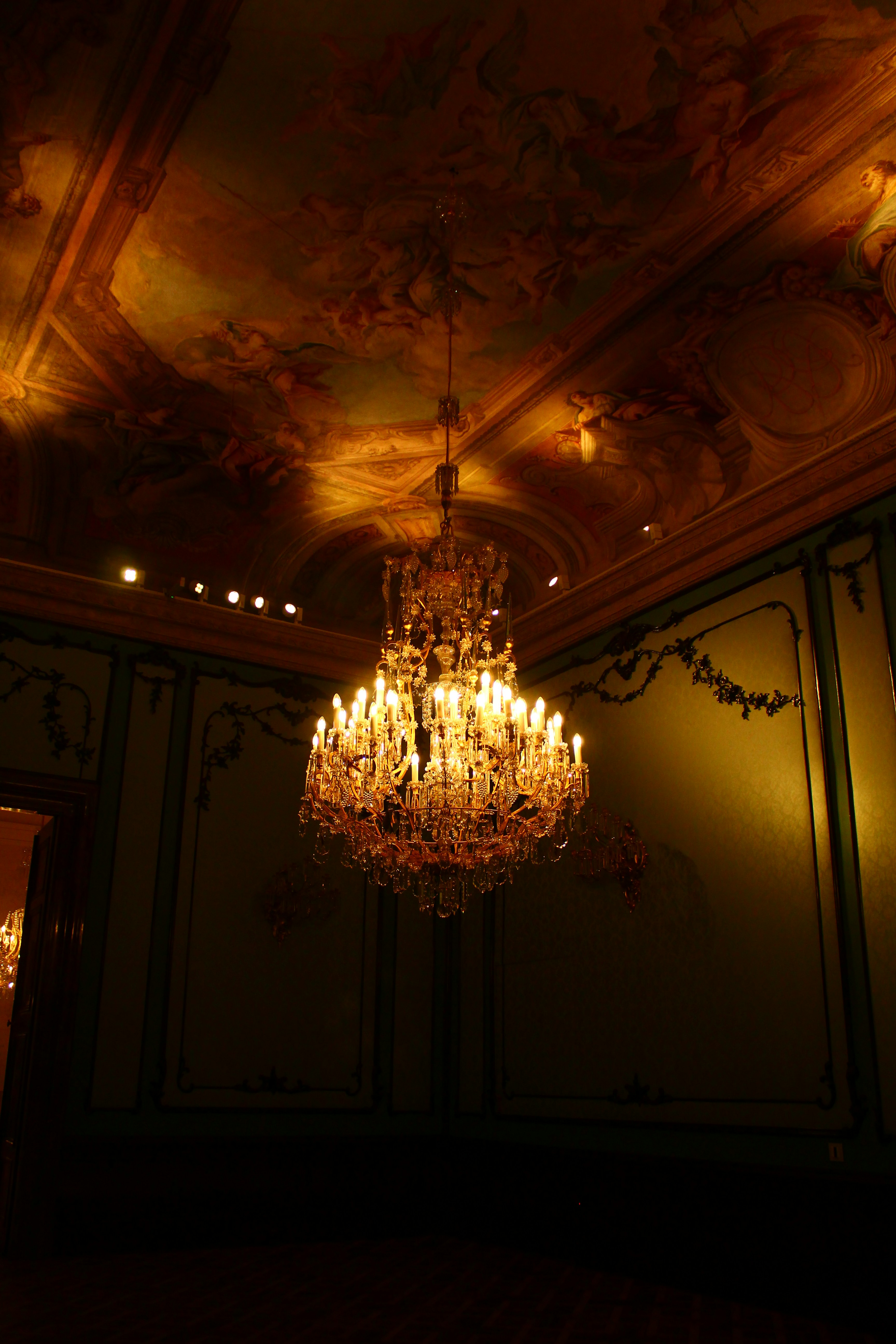 Intricately designed chandelier illuminated in a lavishly decorated room with ornate ceiling details.