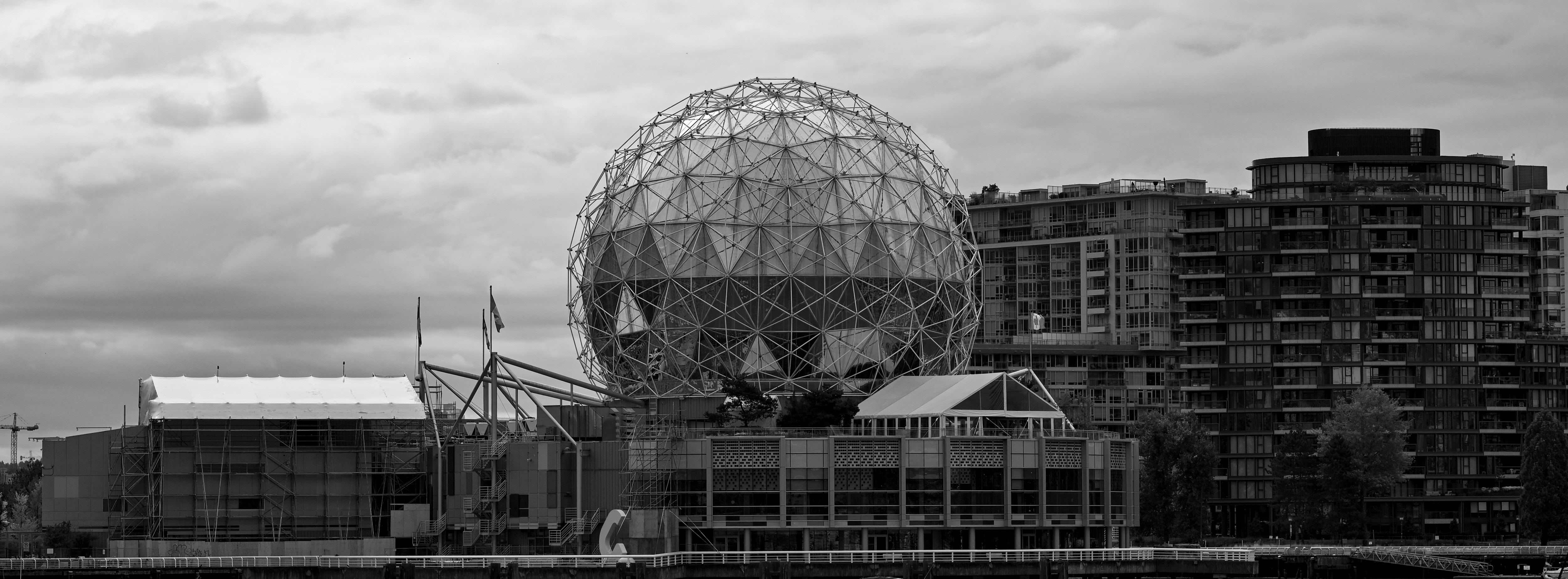 Geodesic dome structure surrounded by modern buildings under a cloudy sky. A blend of architecture and nature in an urban setting.