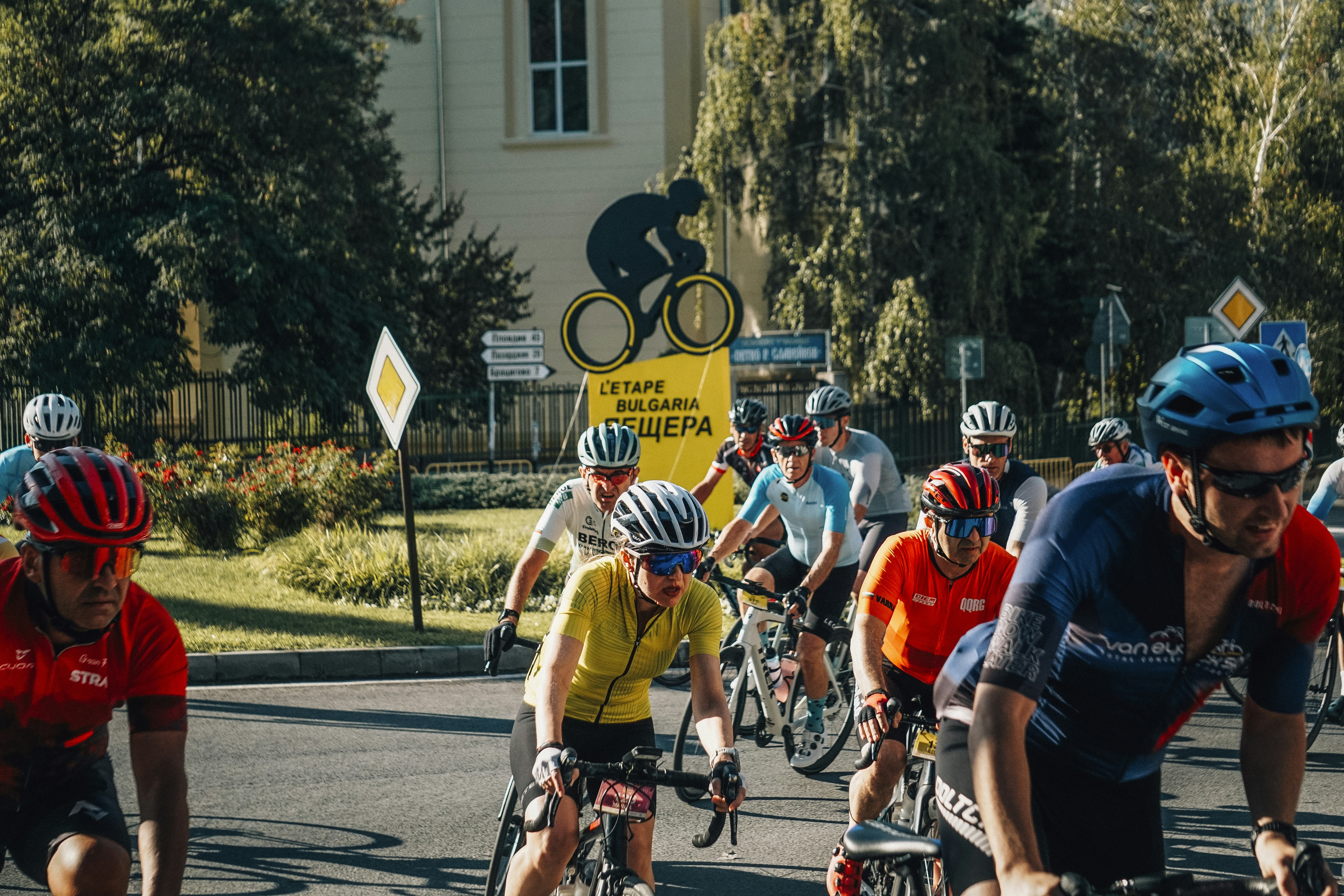 Cyclists racing through a scenic route, with a prominent cycling-themed sign in the background. The scene captures the energy of a competitive event.