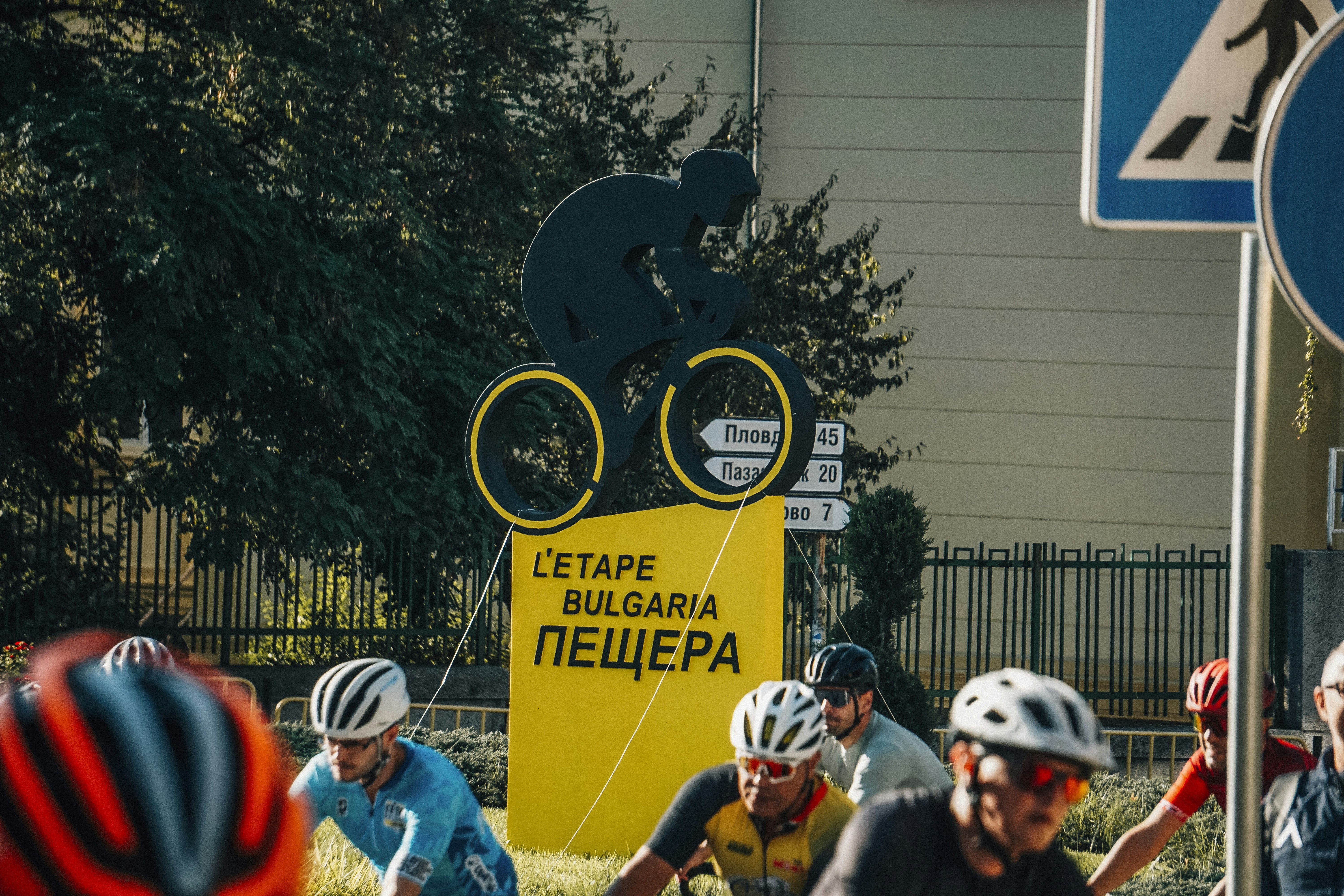 Cyclists in a race pass an urban landmark under a painted cyclist sculpture