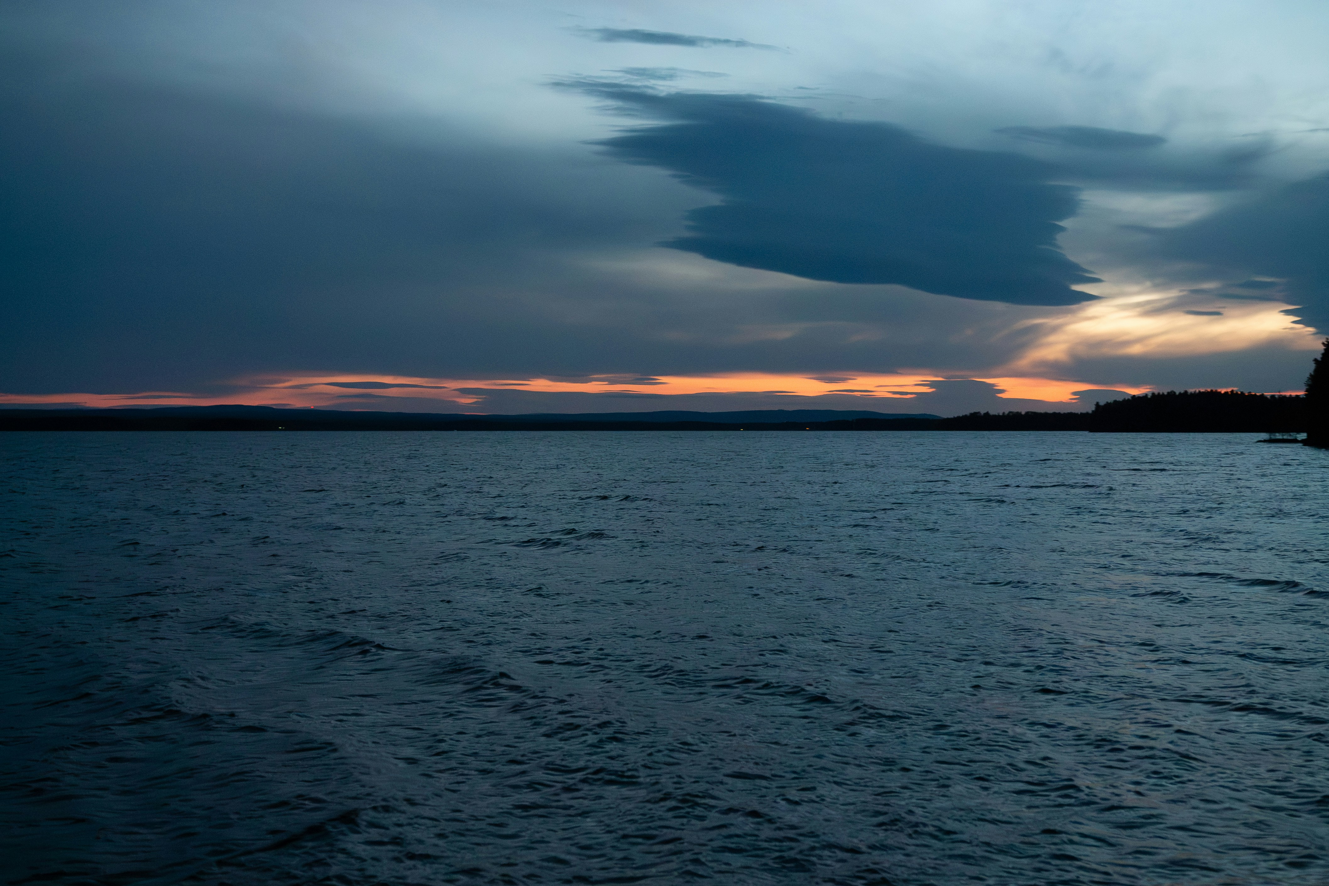 A breathtaking sunset paints the sky with vibrant blue and orange hues, casting a serene glow over the calm water. The dramatic silhouette of a treeline and distant mountains frames this peaceful, twilight landscape. | Dark clouds gather over a calm sea at sunset