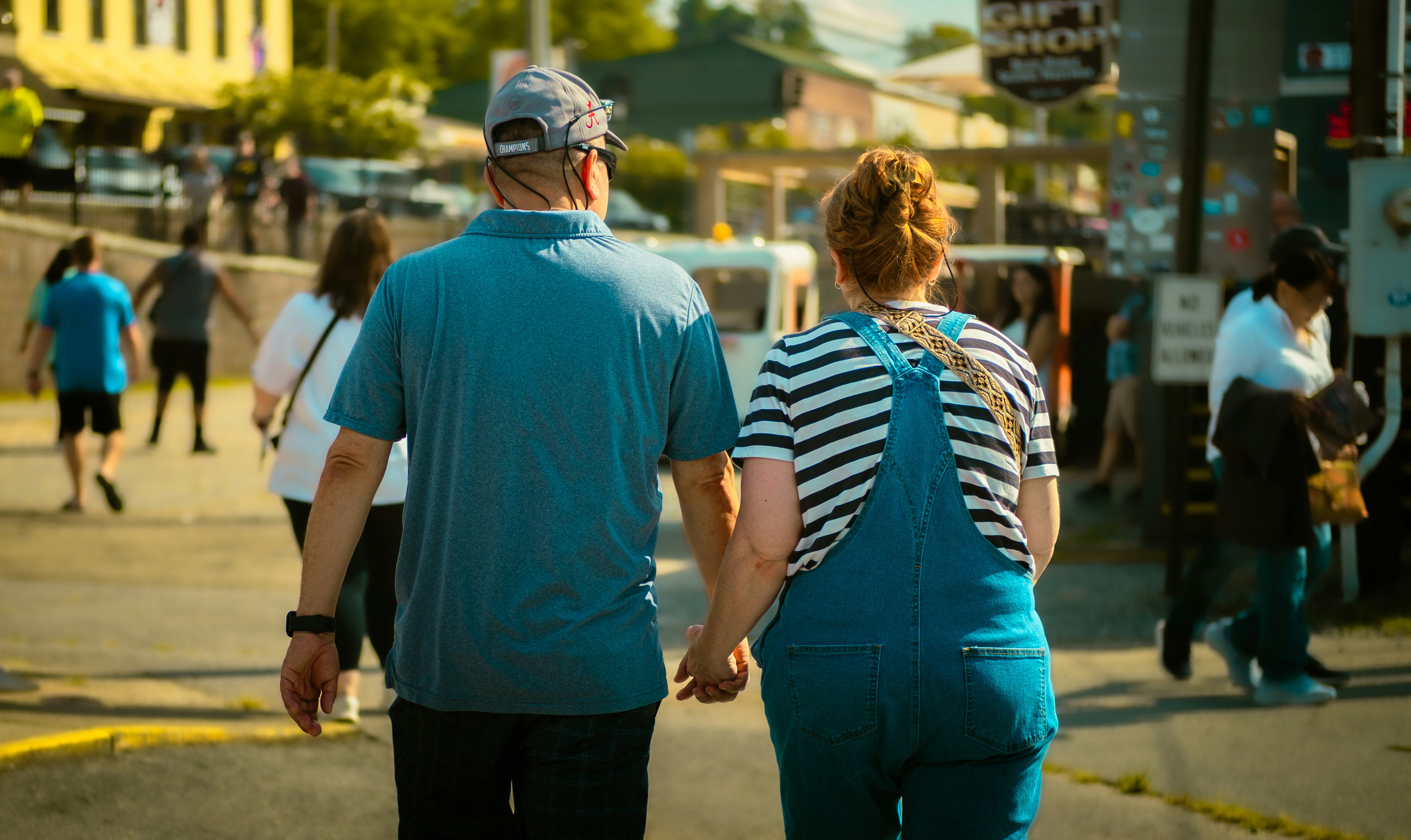 Couple holding hands while walking through a bustling outdoor market, surrounded by a vibrant mix of people. The scene captures a moment of connection amidst the activity.