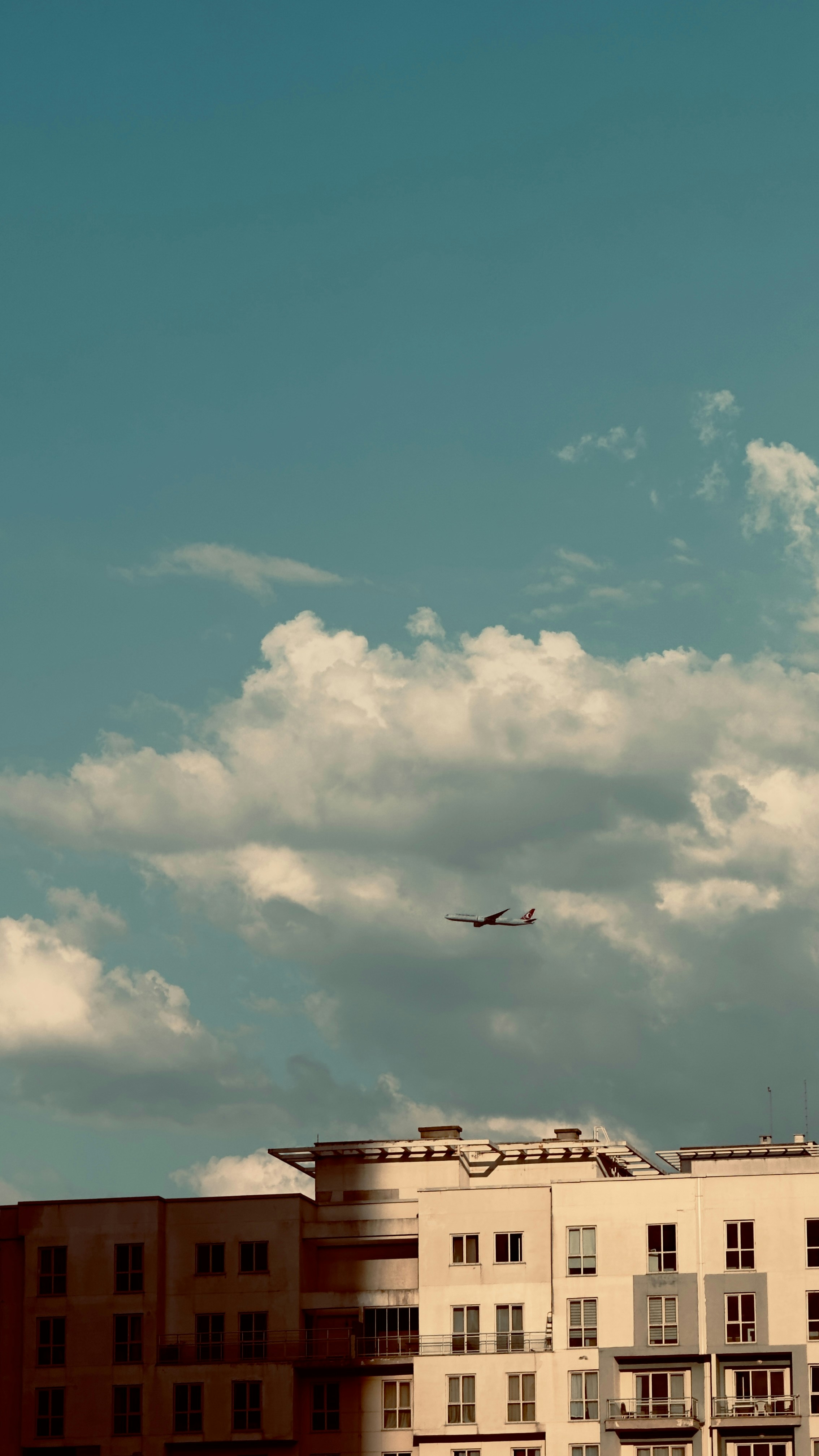 Airplane gliding through a blue sky dotted with fluffy clouds, framed by a modern urban building below.