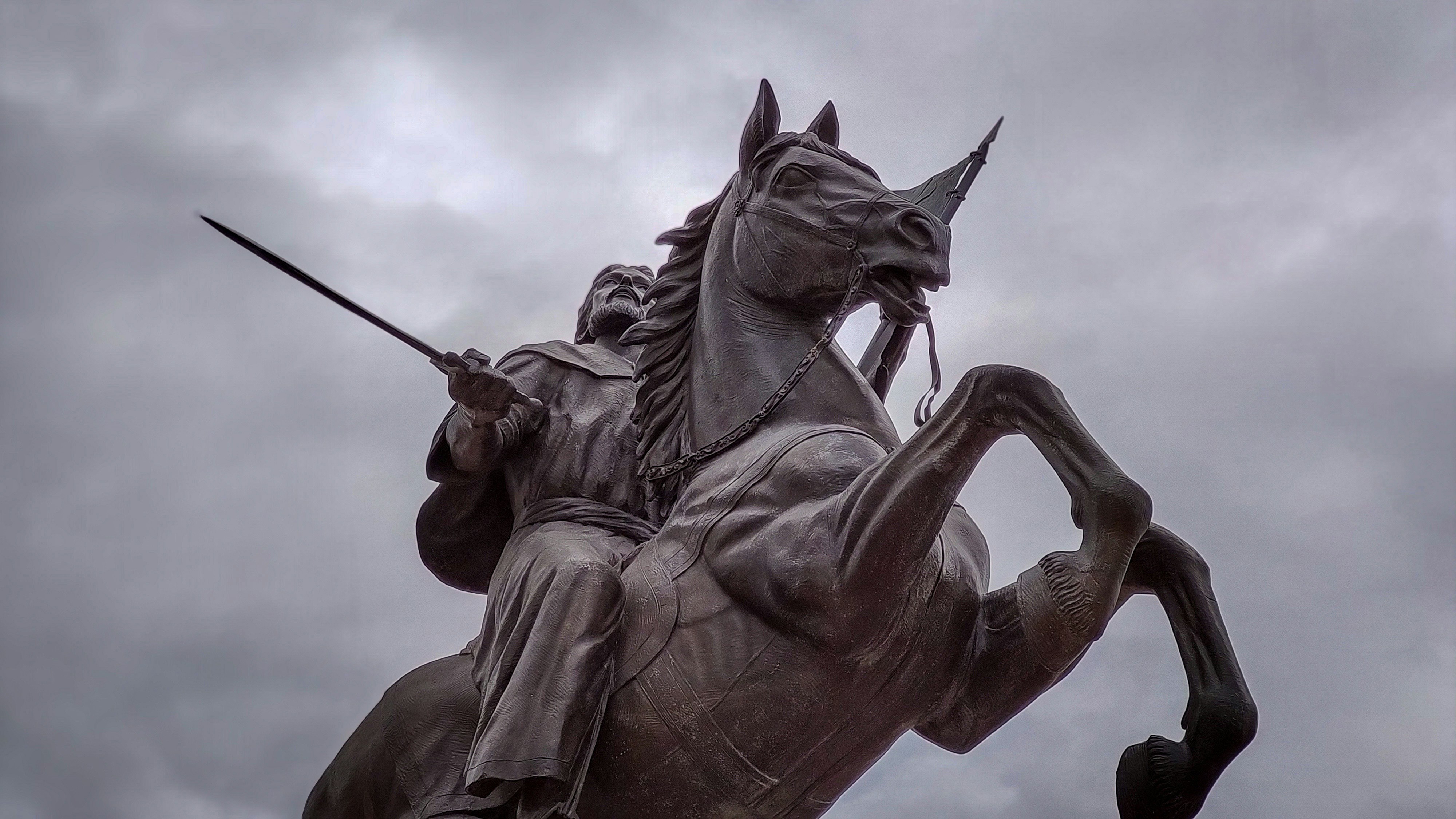 A dynamic bronze statue of a mounted warrior, brandishing a spear, set against a dramatic cloudy sky.