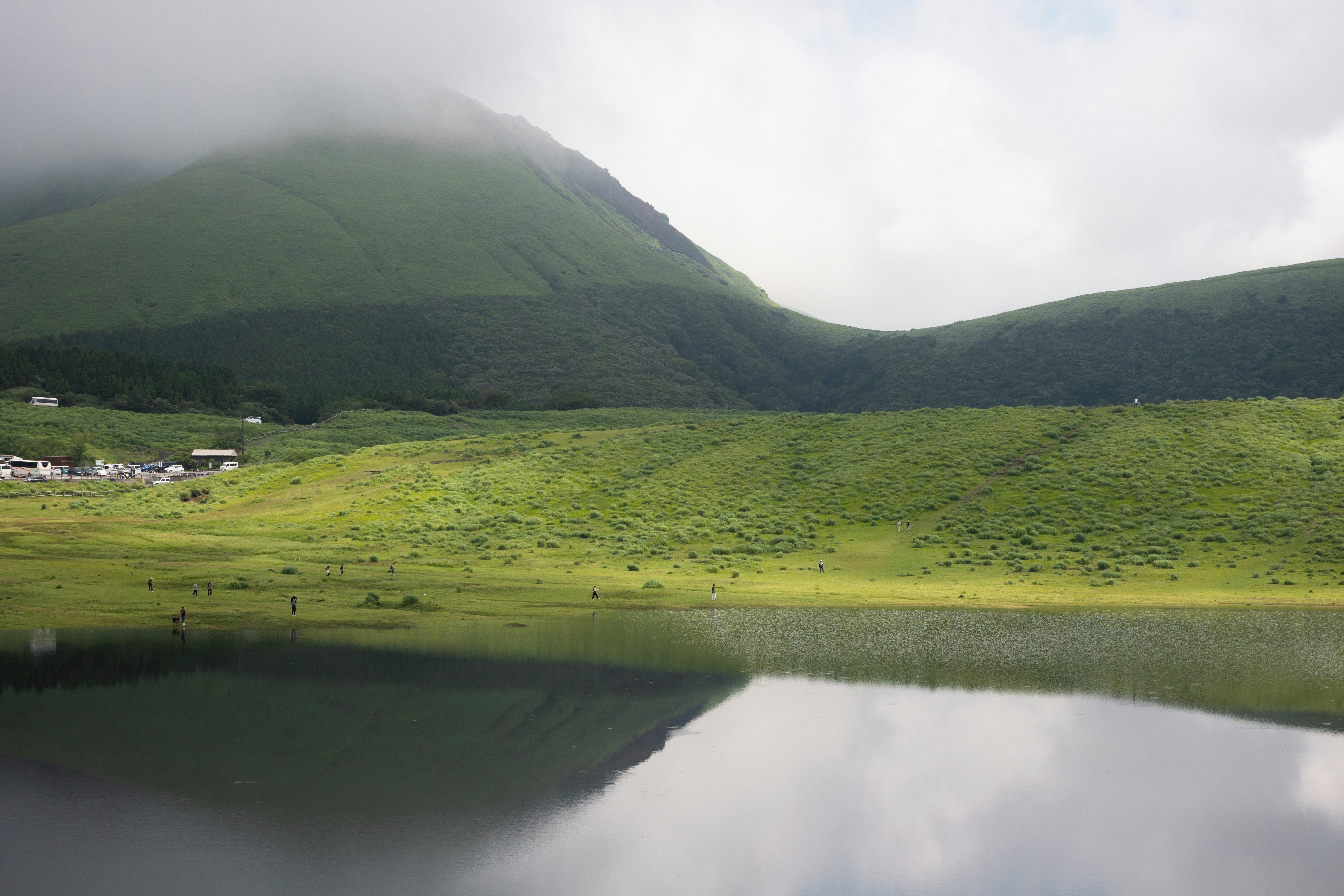 Lush green hills rise gently above a serene lake, reflecting the vibrant landscape under a cloudy sky.