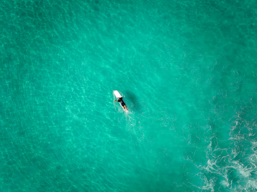 A lone surfer paddles through crystal-clear turquoise waters under a bright sky. The image captures the tranquility and adventure of ocean surfing.