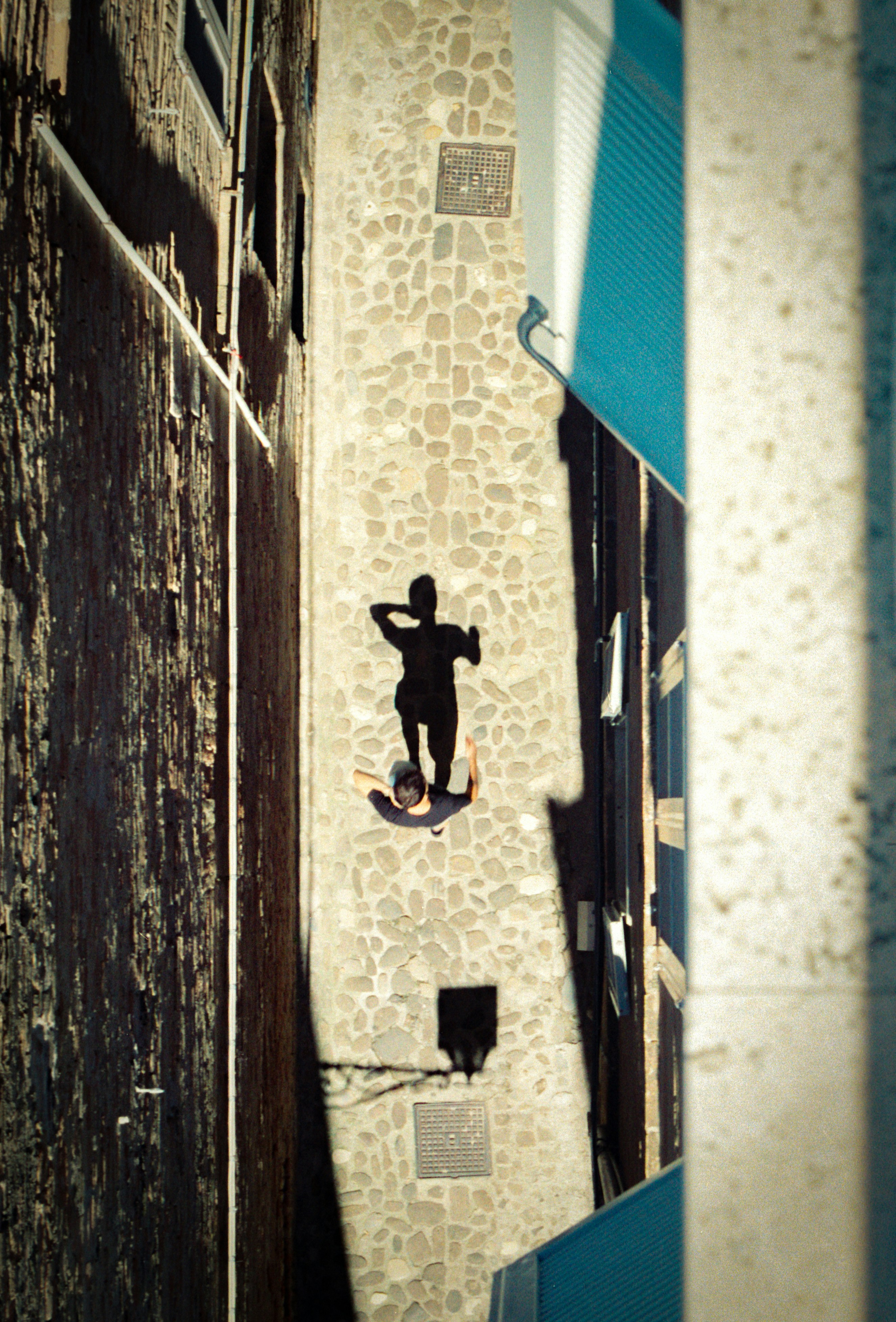 This is one of my favorite photos. From the rooftop terrace, I could see that the sun would soon be at just the right angle to shine straight down the alley. All I had to do then was wait for someone photogenic enough to walk into the scene. It’s amazing, by the way, how fast the sun seems to move when you really wish it wouldn’t. With a digital camera, I would’ve just taken two hundred shots and picked the best one afterwards. Shooting on film is a bit trickier, so I had to be confident enough to settle for just three frames. One ended up overexposed, another had an awkward shadow... | Shadow of a person dancing on a cobblestone street