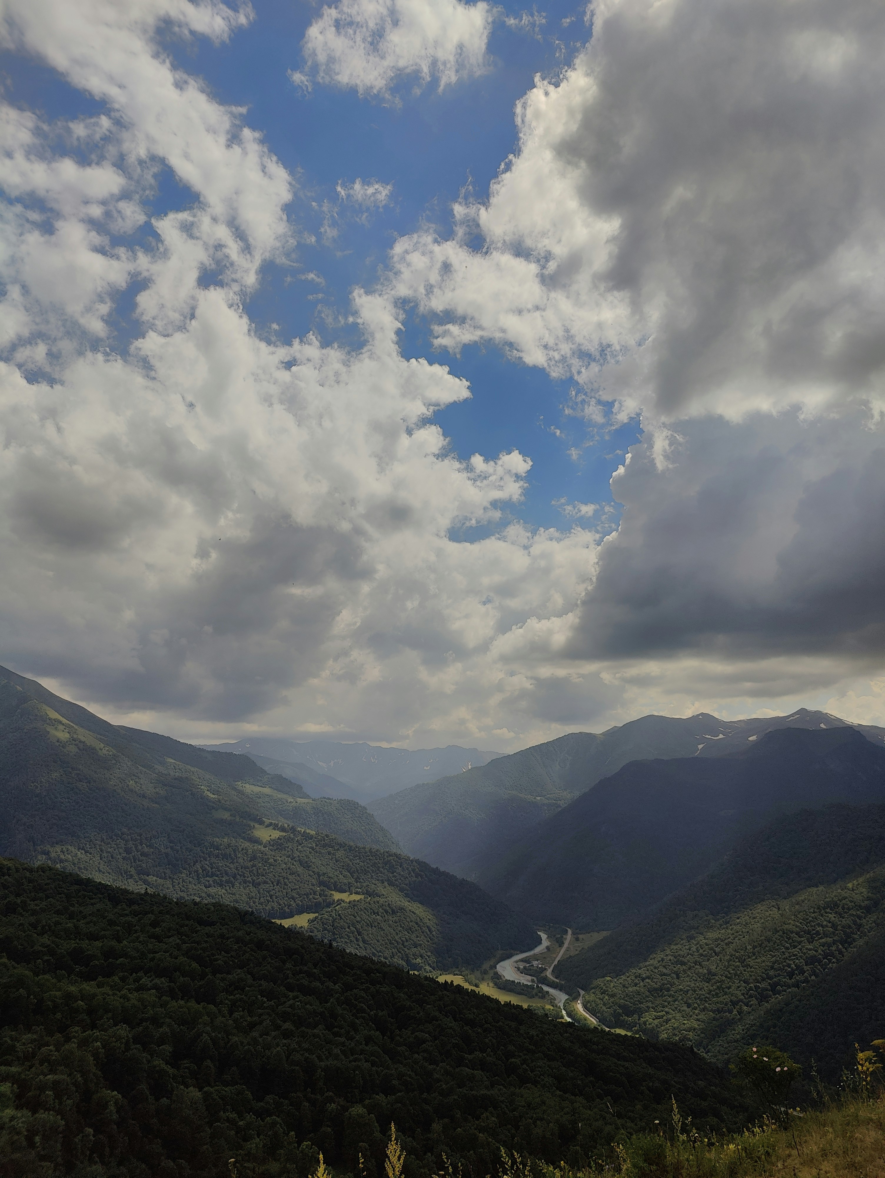 Lush green mountains cradle a winding river beneath a dramatic sky filled with clouds. The scene captures the tranquil beauty of nature's contours.