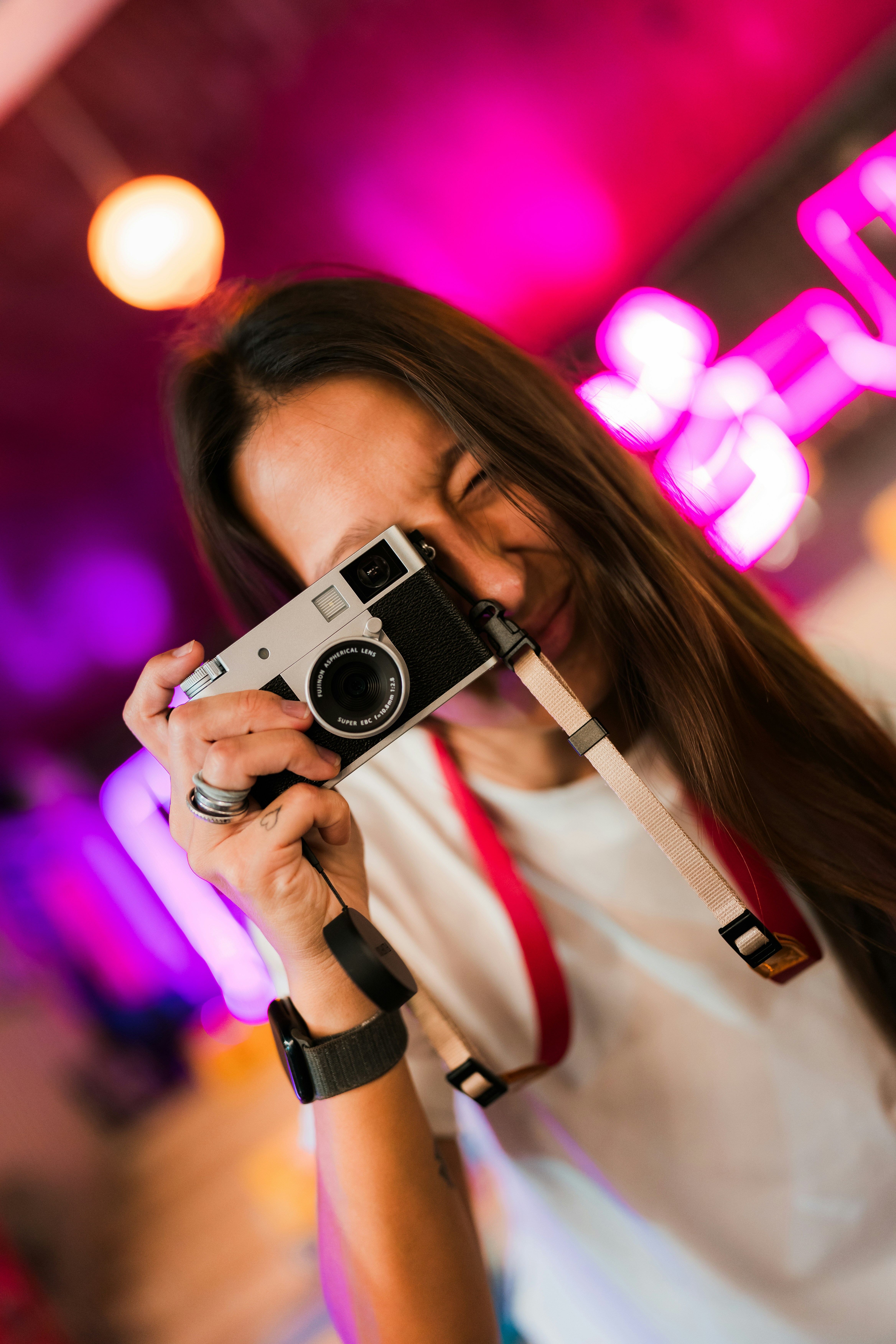 A young woman playfully captures a moment with a vintage camera, surrounded by vibrant neon lights in a lively setting.
