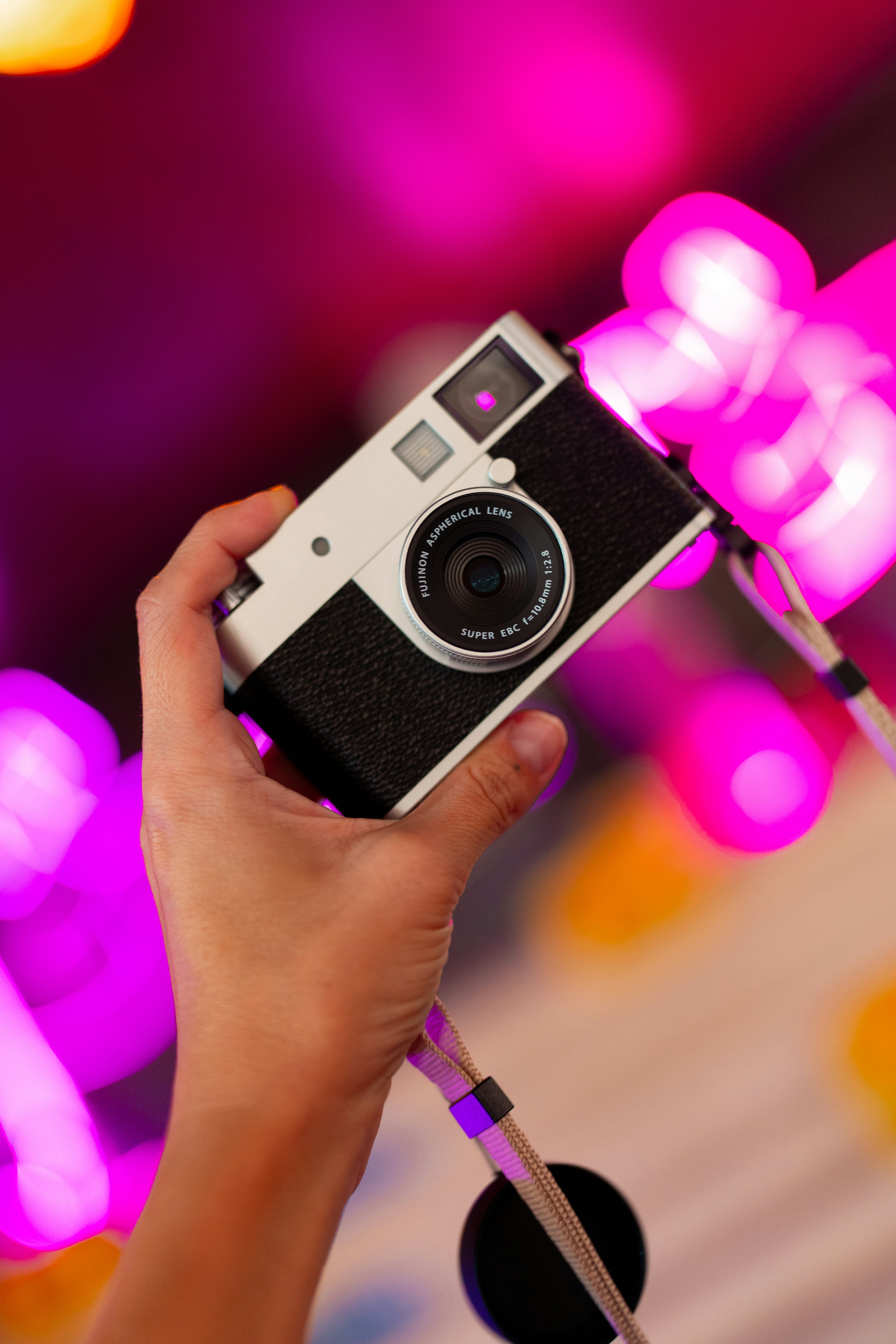 Close-up of a hand holding a Fujifilm X-HALF digital camera against a vibrant, colorful bokeh background in pink, purple, and orange hues. The silver and black retro-style body and compact prime lens stand out, combining vintage design with modern technology. | Hand holding a vintage camera with bokeh lights