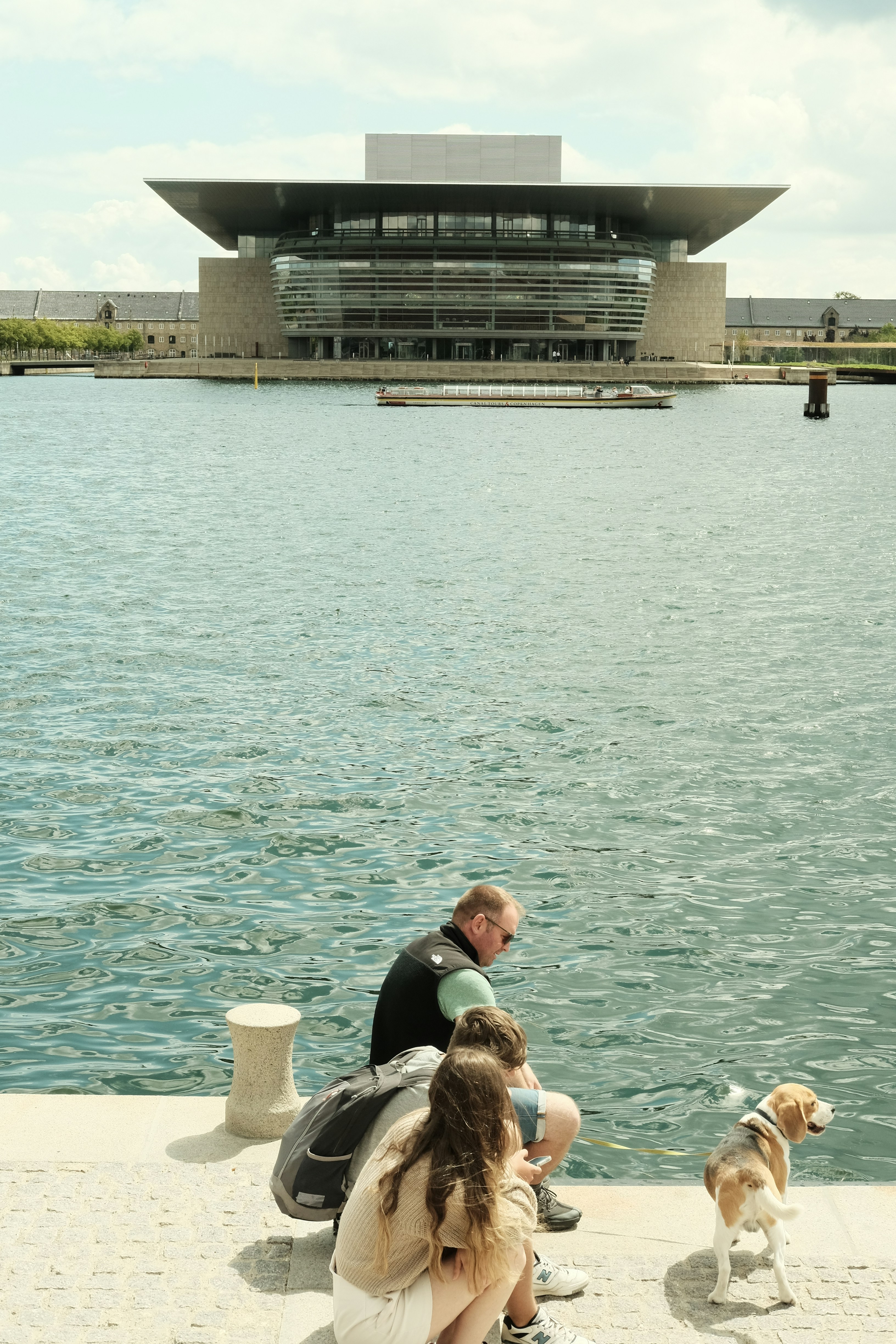 People with a dog sit along a waterfront beside a contemporary building, the scene framed by calm water and a wide sky.