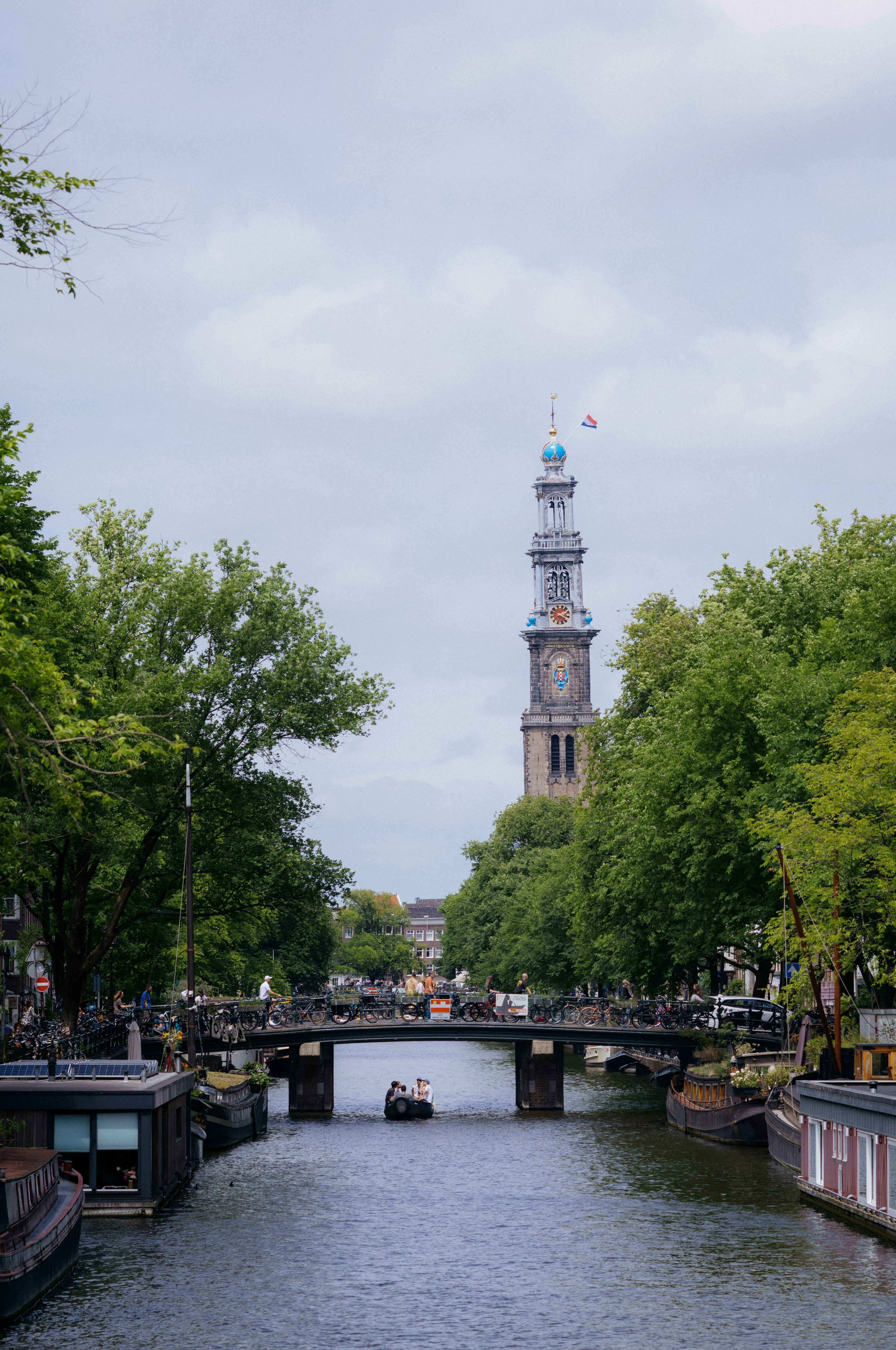 A serene Amsterdam canal scene featuring a bridge and the iconic Westerkerk tower in the background, surrounded by lush greenery.