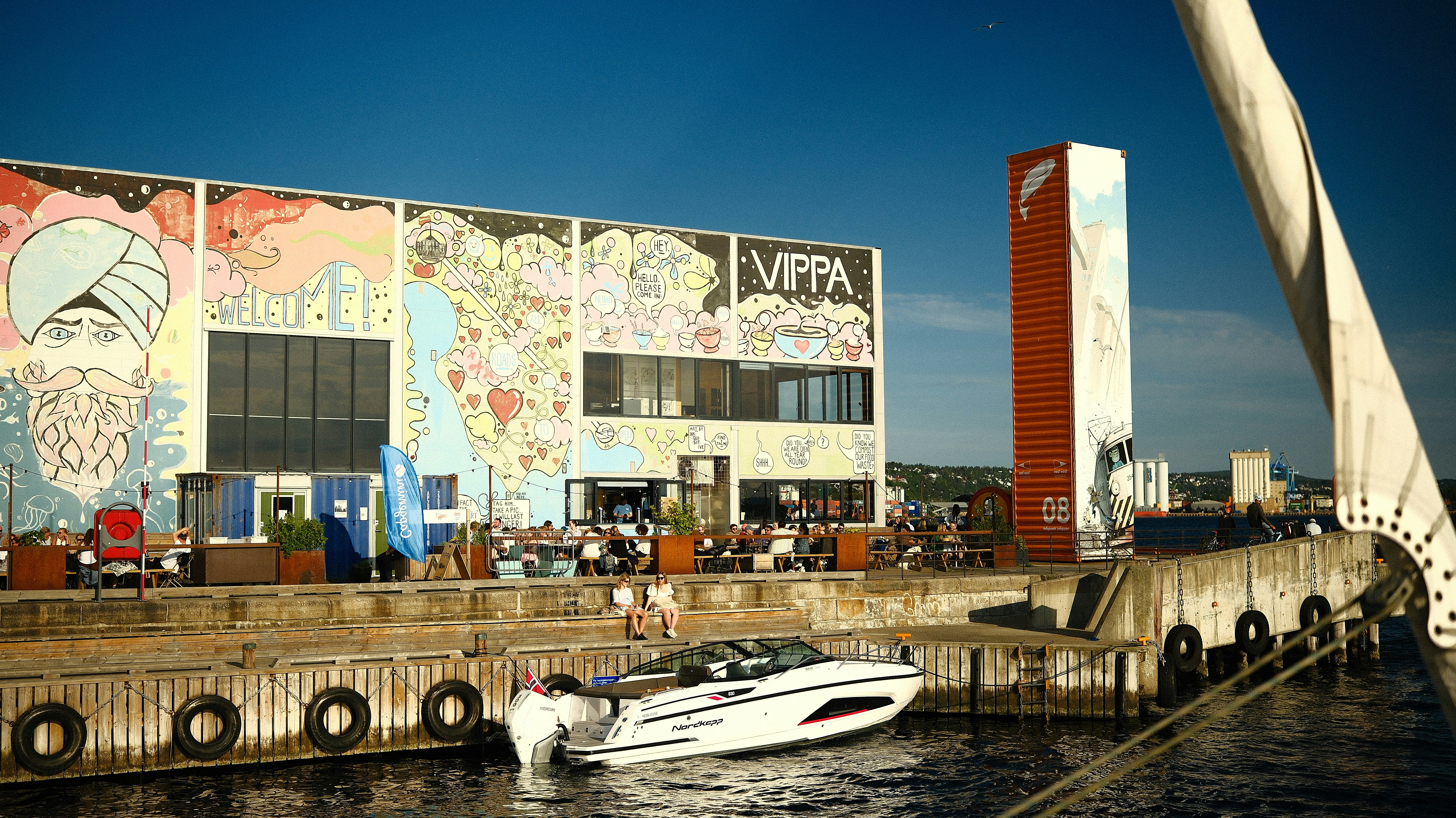 Summer afternoon at Vippa food hall on Akershusstranda, Oslo: a vibrant waterfront scene with a colourful “WELCOME” mural, people relaxing and enjoying drinks on a wooden pier, and a small boat gently bobbing in the harbour beneath a deep-blue sky. Warm, lively and cinematic — ideal as a travel or lifestyle background for website headers, banners, social posts, or phone wallpapers. Location: Vippa, Akershusstranda 25, 0150 Oslo, Norway. | Modern building with mural art by a harbor with boat.