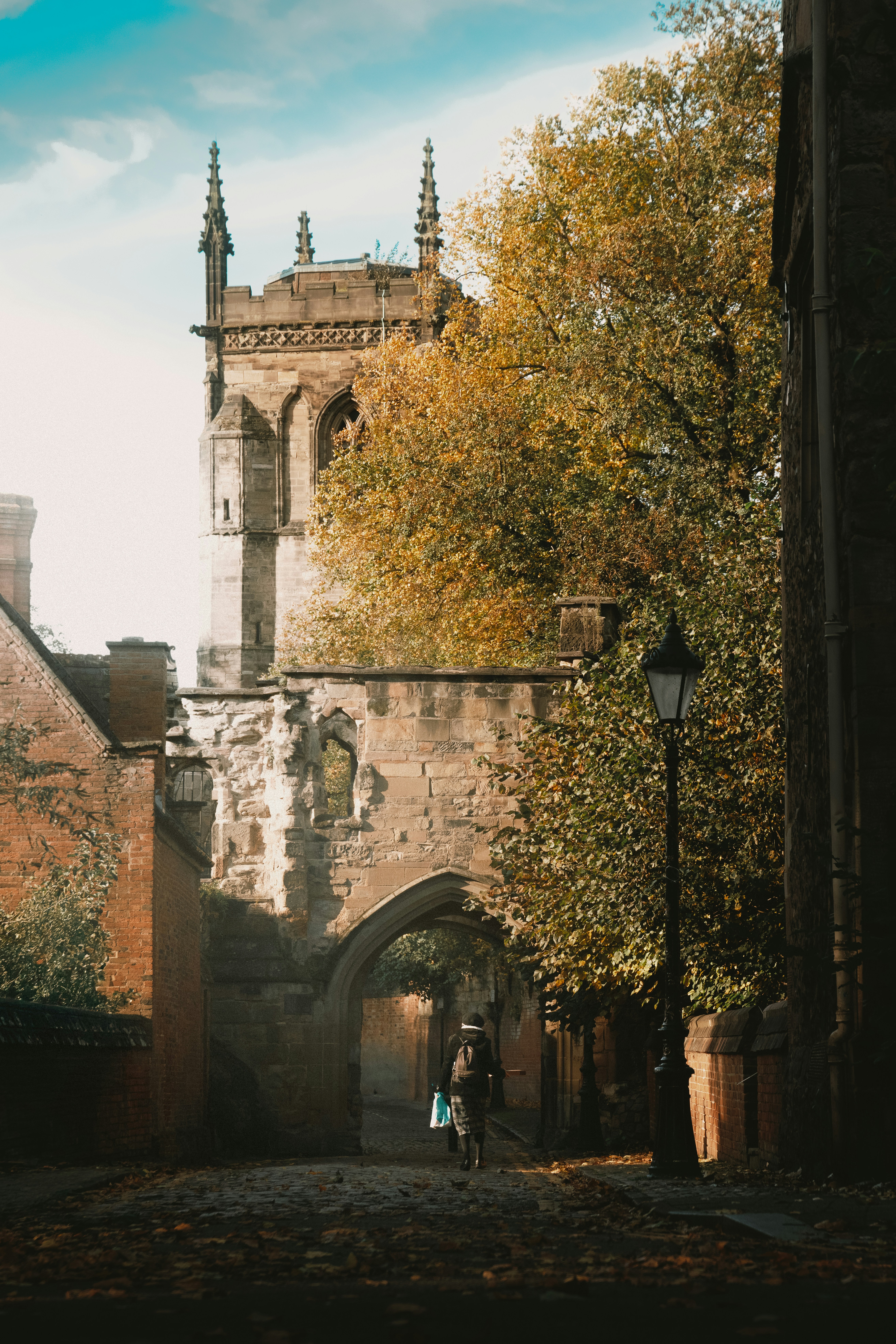 A solitary figure walks beneath an ancient archway, framed by vibrant autumn foliage and a historic tower. The scene evokes a sense of nostalgia and tranquility.