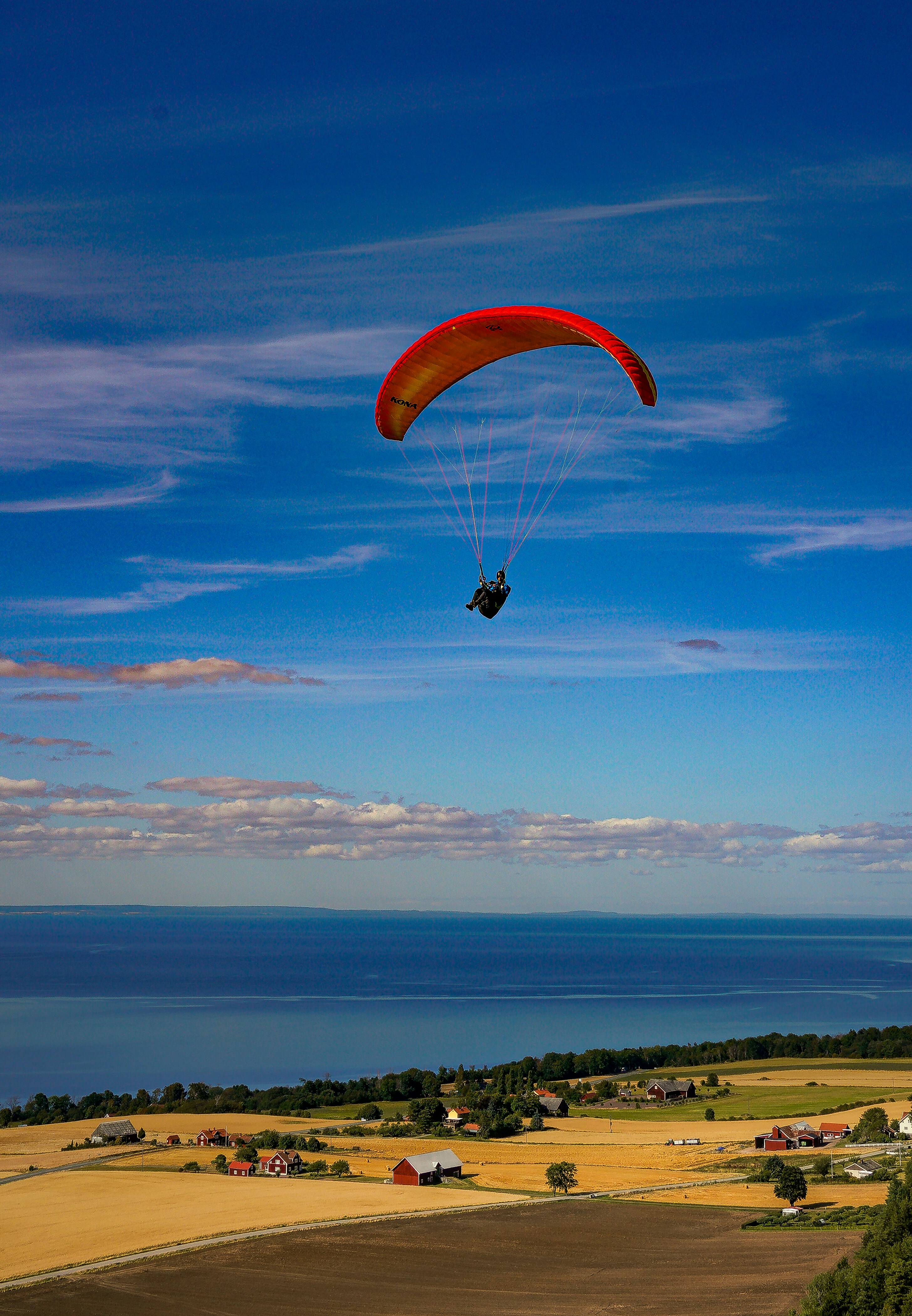 A paraglider gracefully navigates the sky above a patchwork of farmland and a serene lake, showcasing the beauty of aerial adventure.