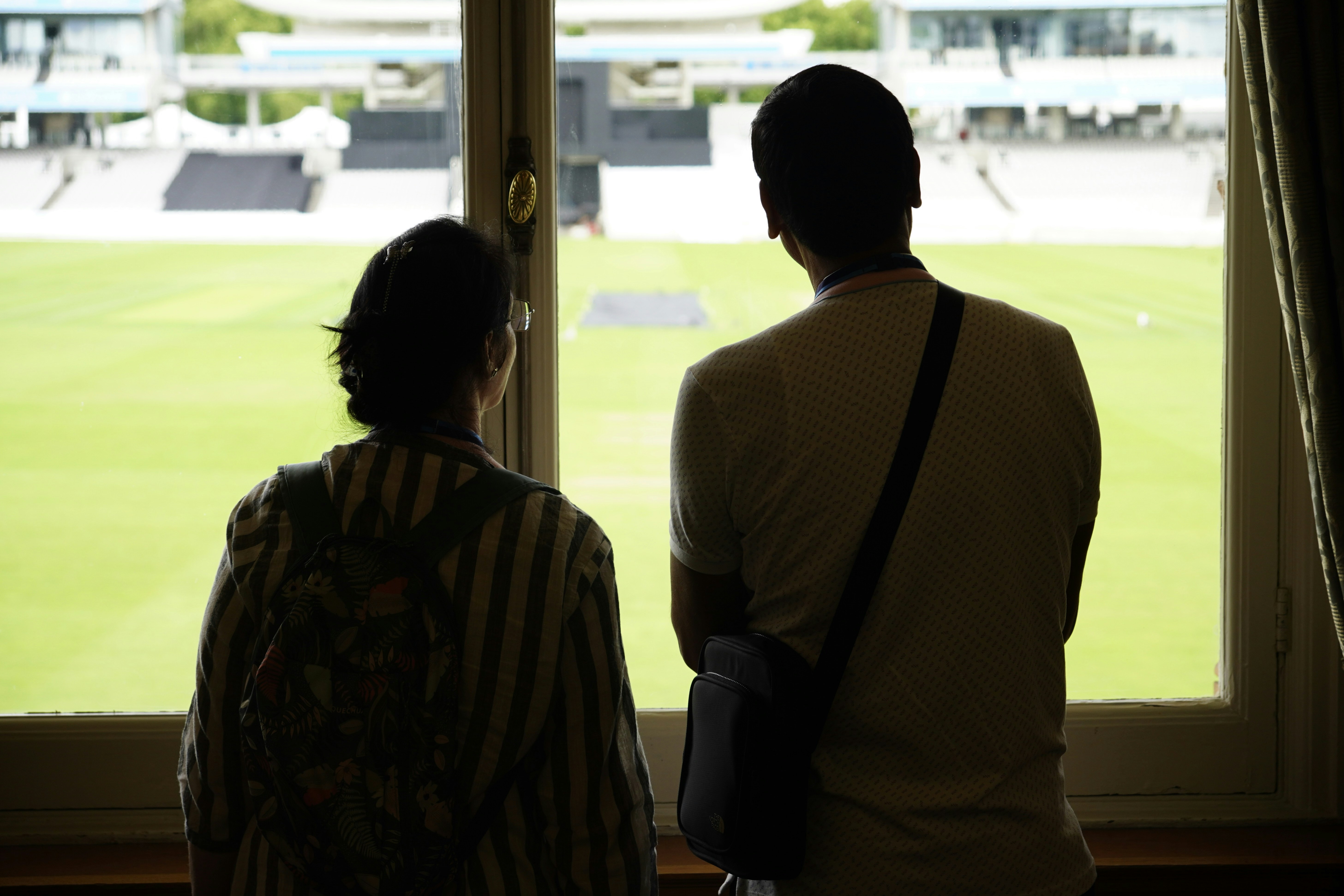 Two spectators gazing out over a cricket field from a window, immersed in the atmosphere of the game.