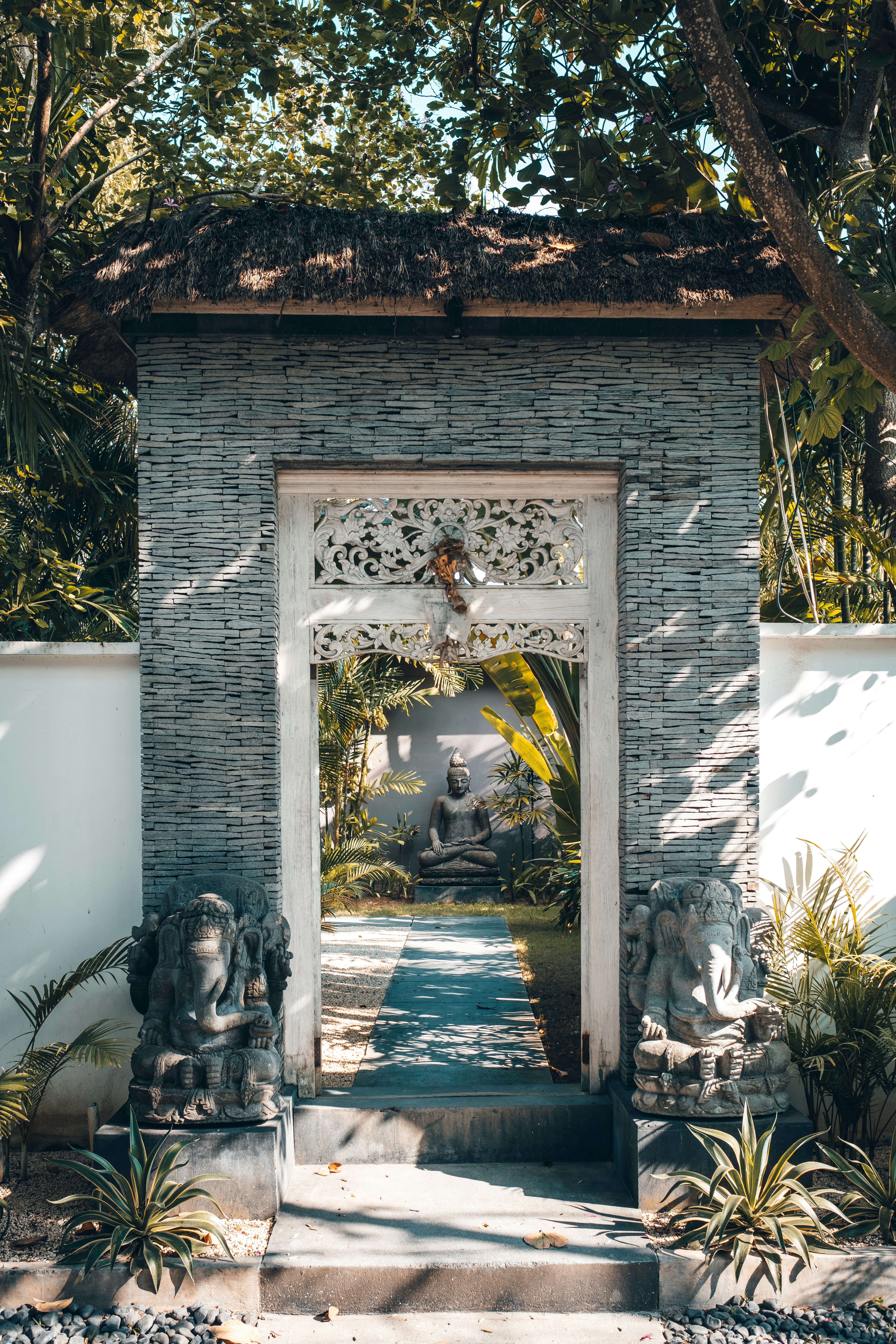 Intricate stone archway flanked by guardian sculptures leads to a peaceful garden featuring a seated Buddha statue. 