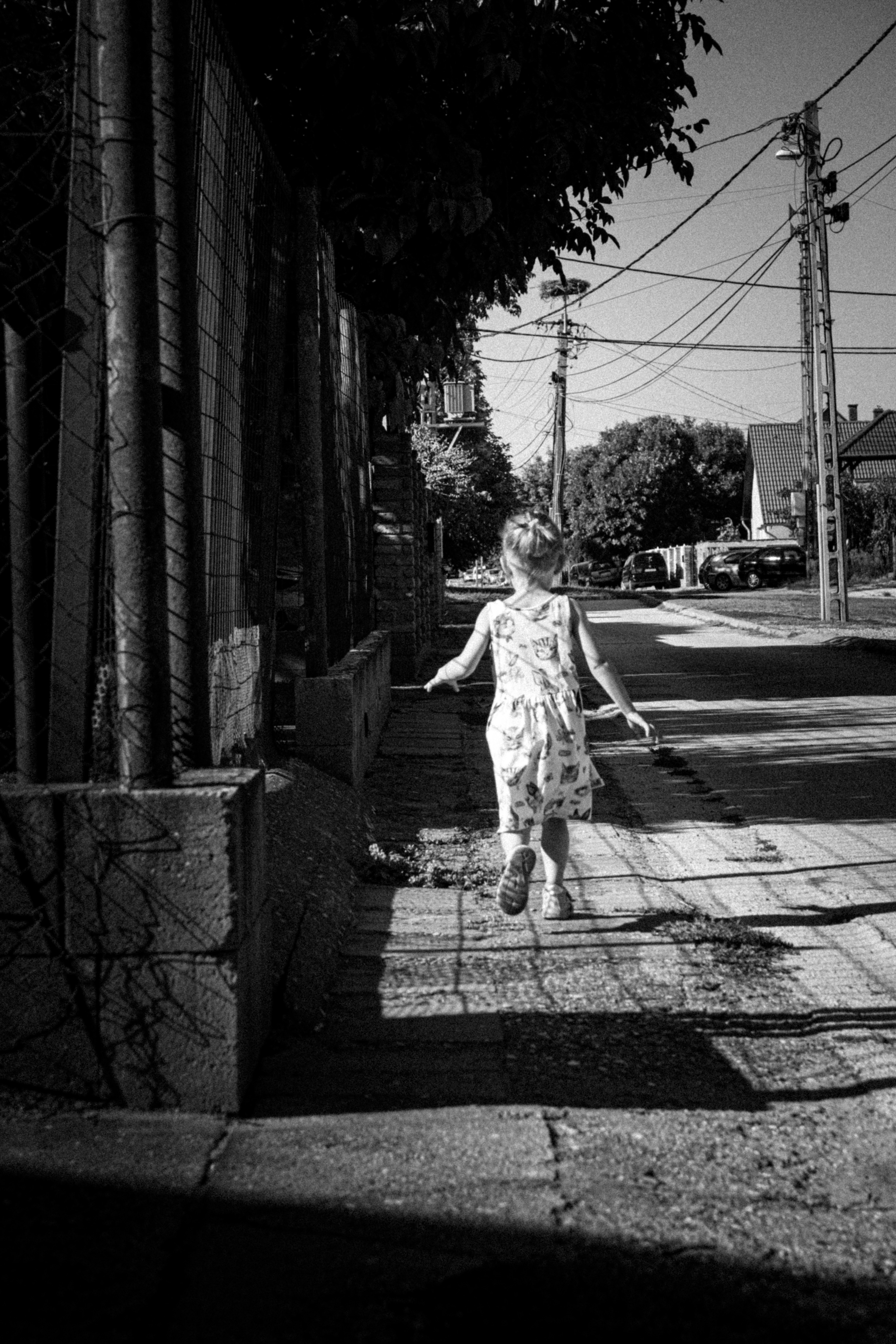 Young girl in a floral dress runs along a sunlit street, framed by shadows and urban elements. The scene captures a moment of playful exploration.