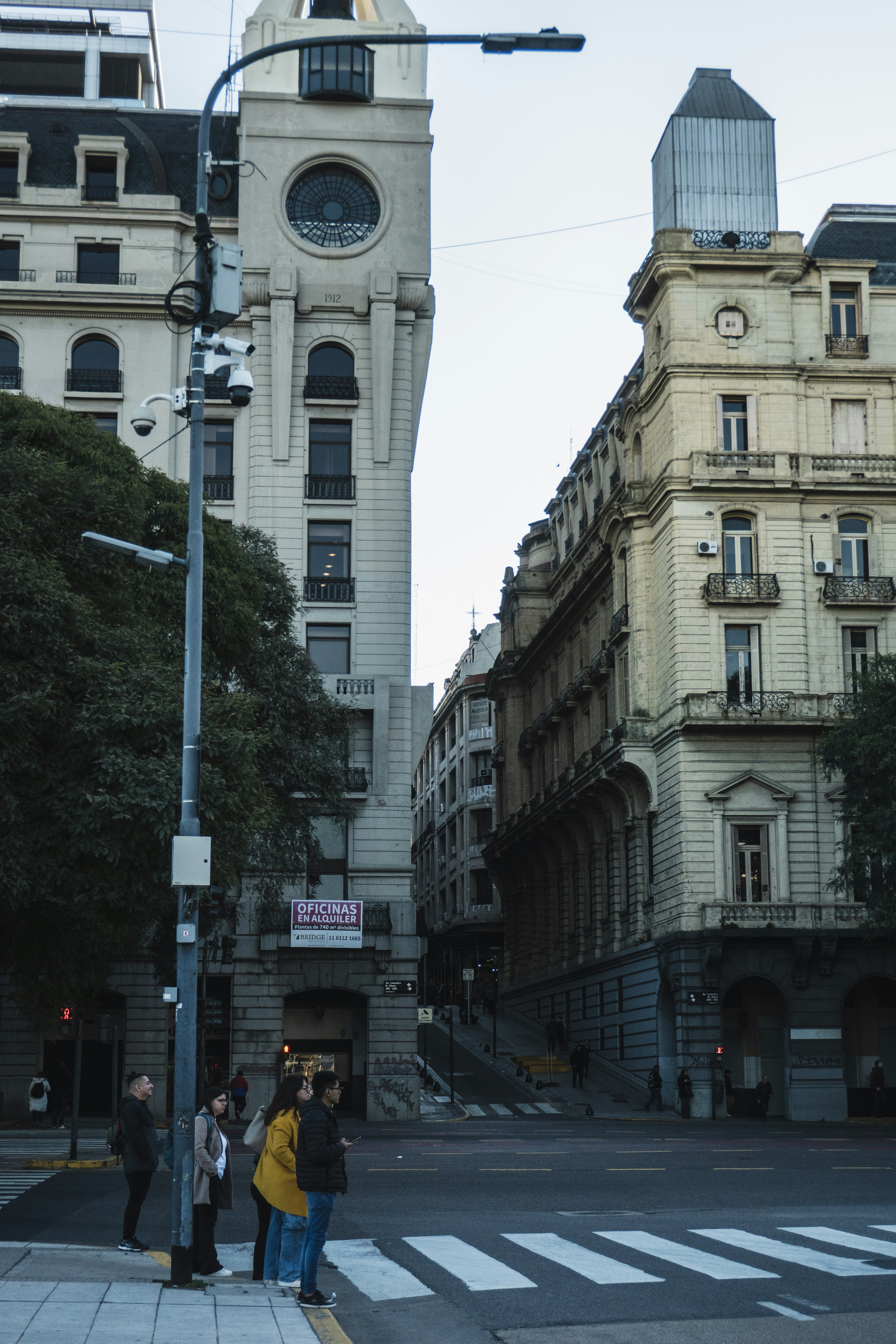 People waiting at a crosswalk in a historic urban setting, flanked by contrasting architectural styles. The scene captures the essence of city life.