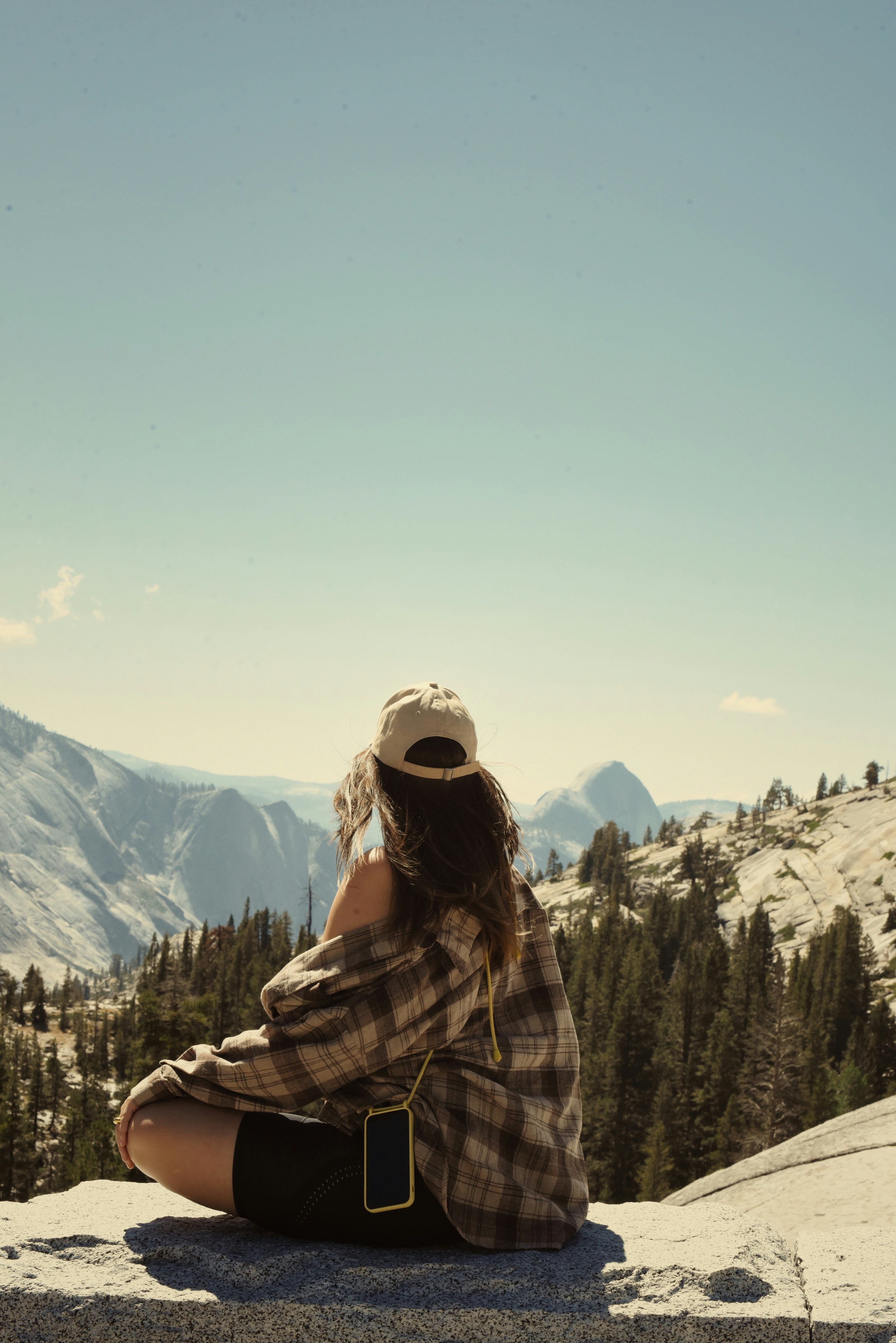 Individual seated on a rock, gazing at a vast mountainous landscape under a clear blue sky. The scene captures a moment of tranquility amidst nature.
