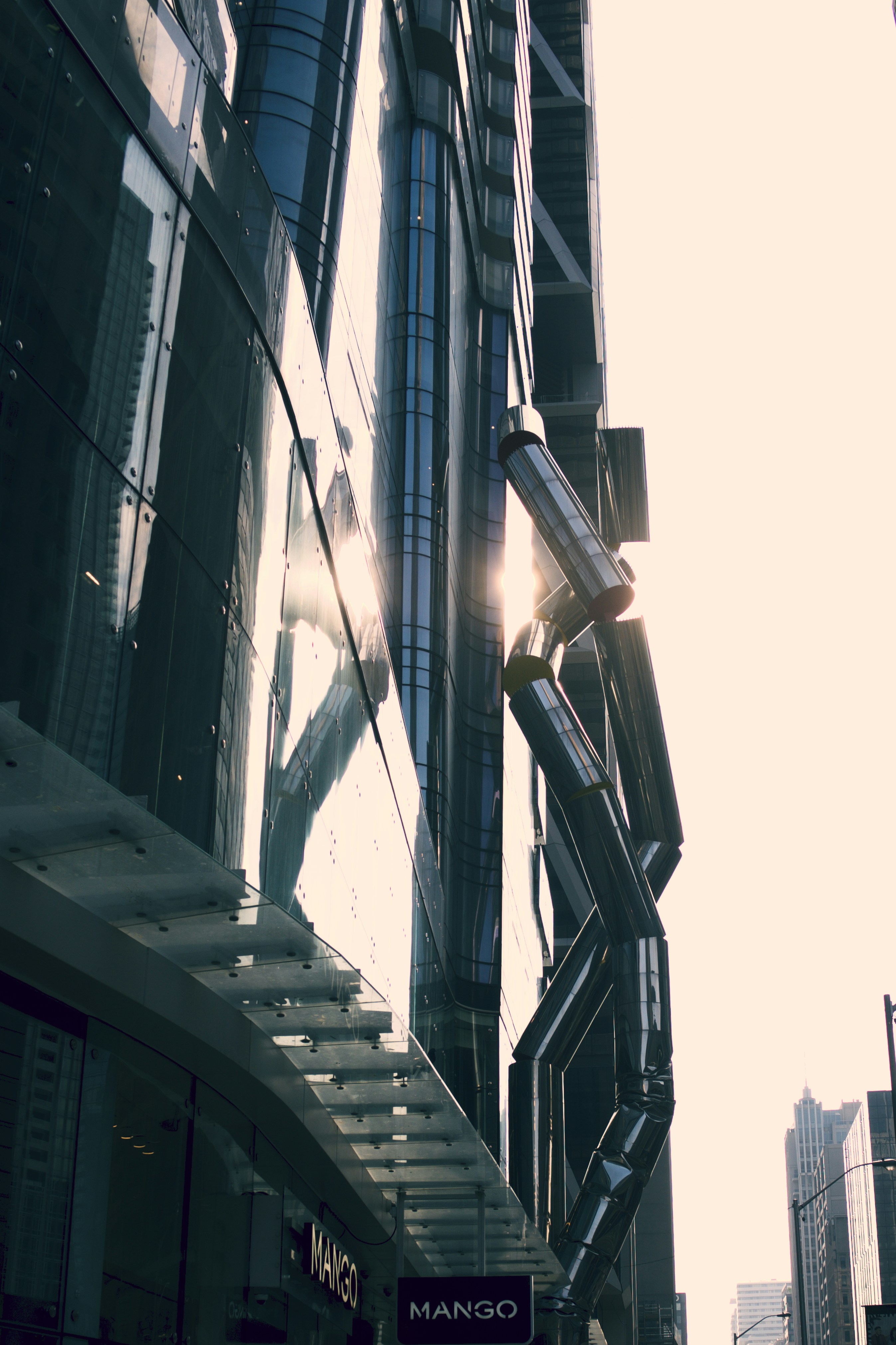 Sleek glass facade of a modern building reflecting sunlight, with a prominent metallic structure accentuating the design. Logo of 'MANGO' visible at the bottom.