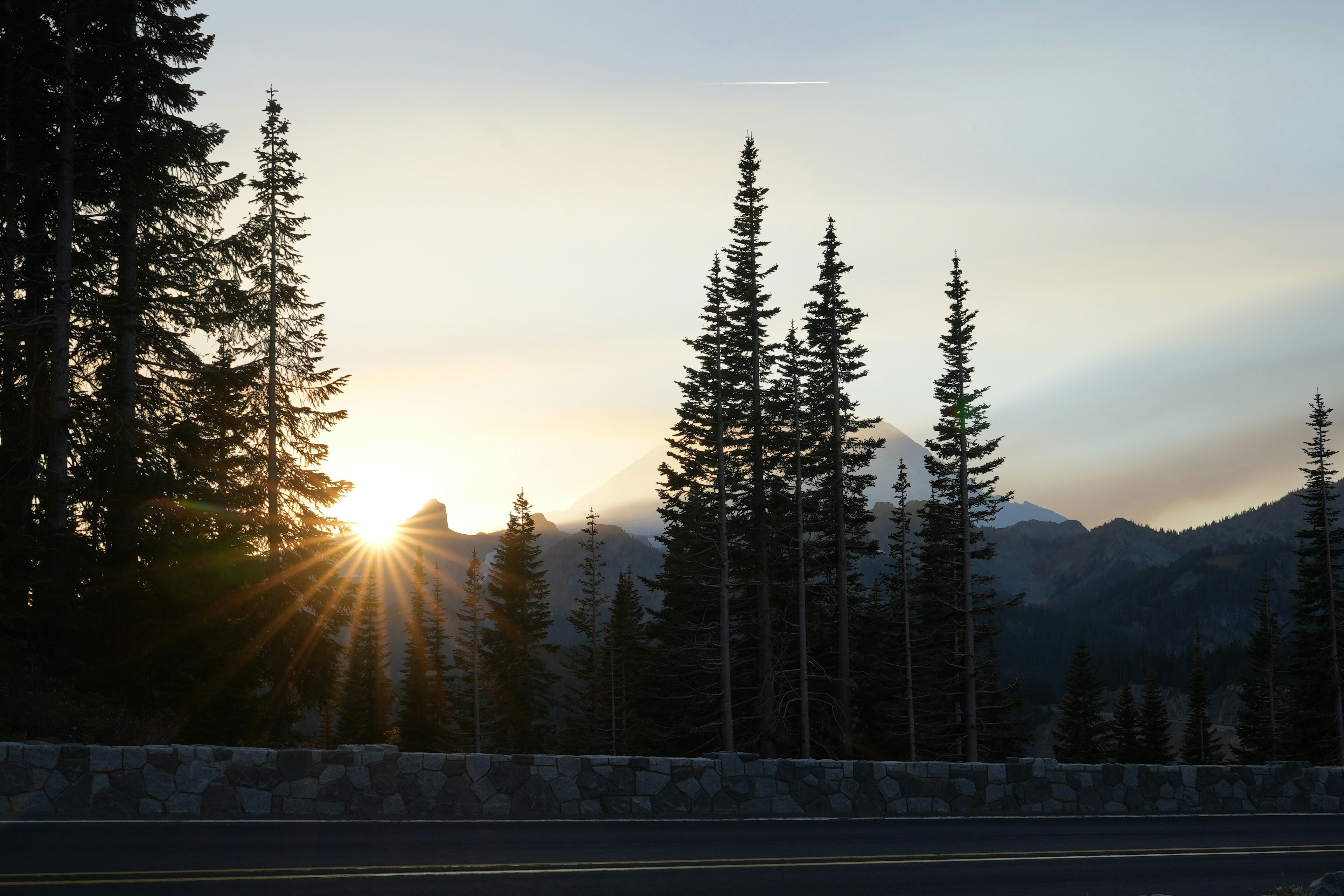 Sunset peeking through tall conifer trees, casting rays over a distant mountain range. The scene captures the serene transition from day to night.
