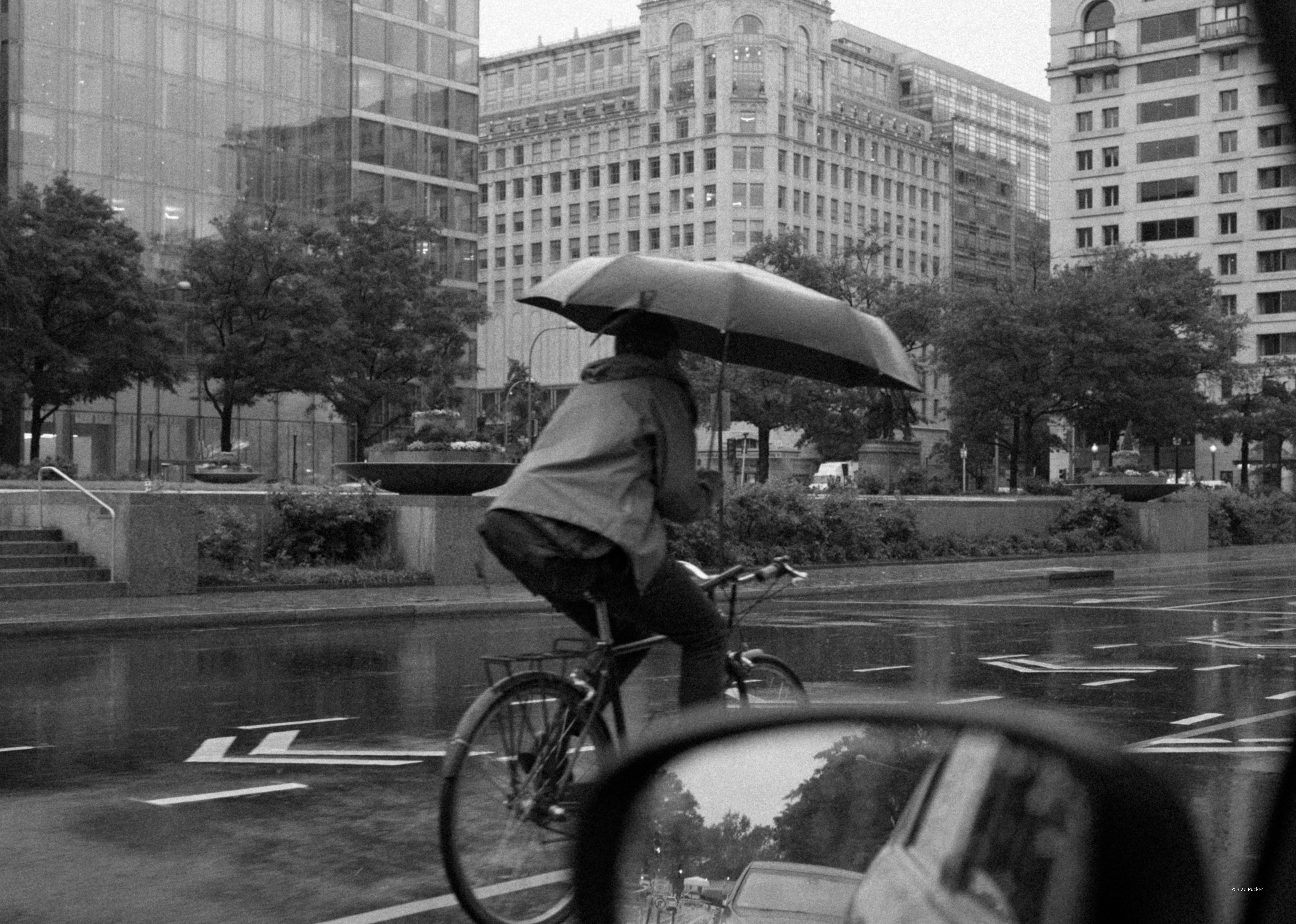 A cyclist navigates a rainy cityscape, shielded by an umbrella, as reflections dance on wet pavement.