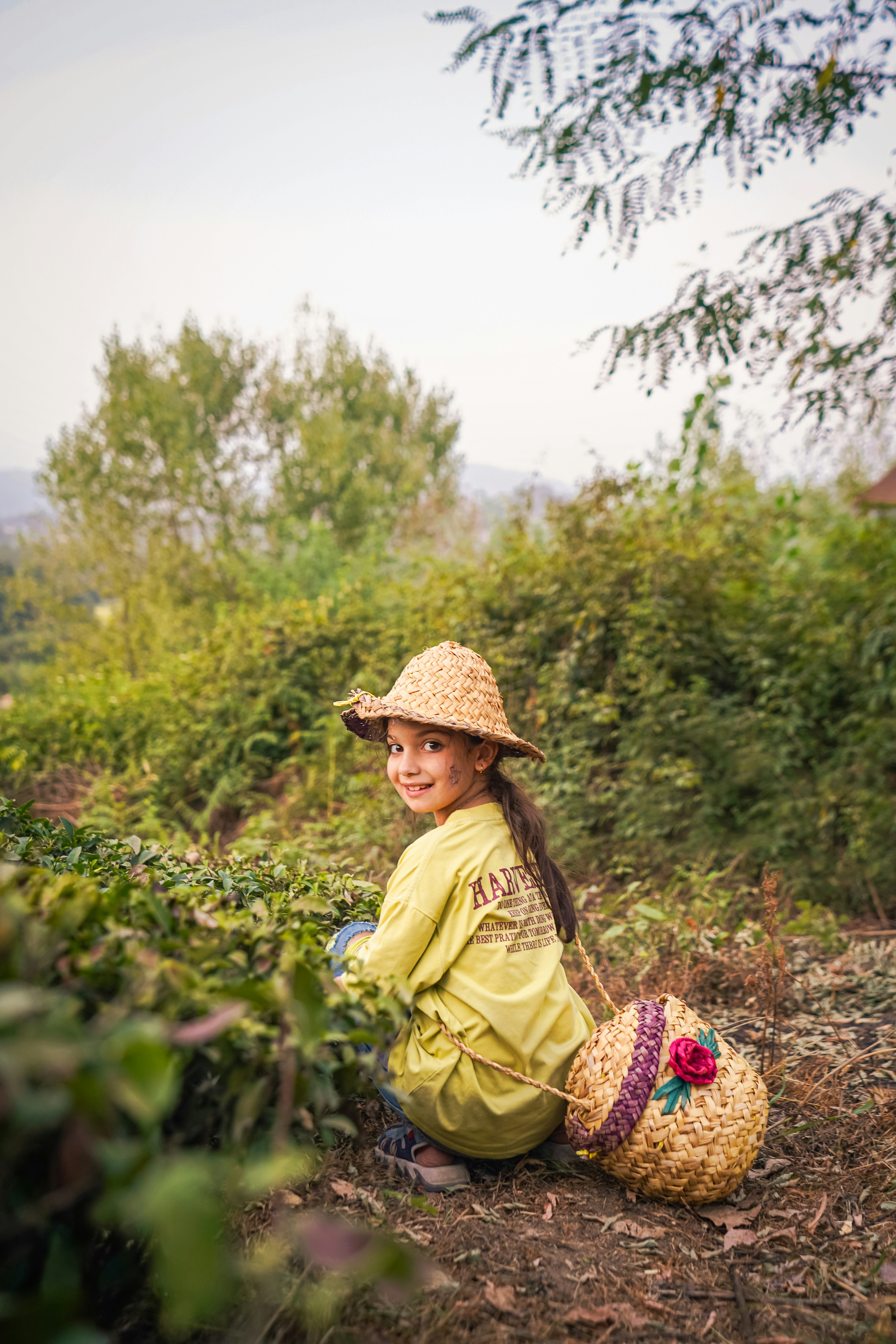 Young girl in a straw hat tending to a garden, smiling while holding a decorative basket. Lush greenery surrounds her, capturing the essence of rural life.