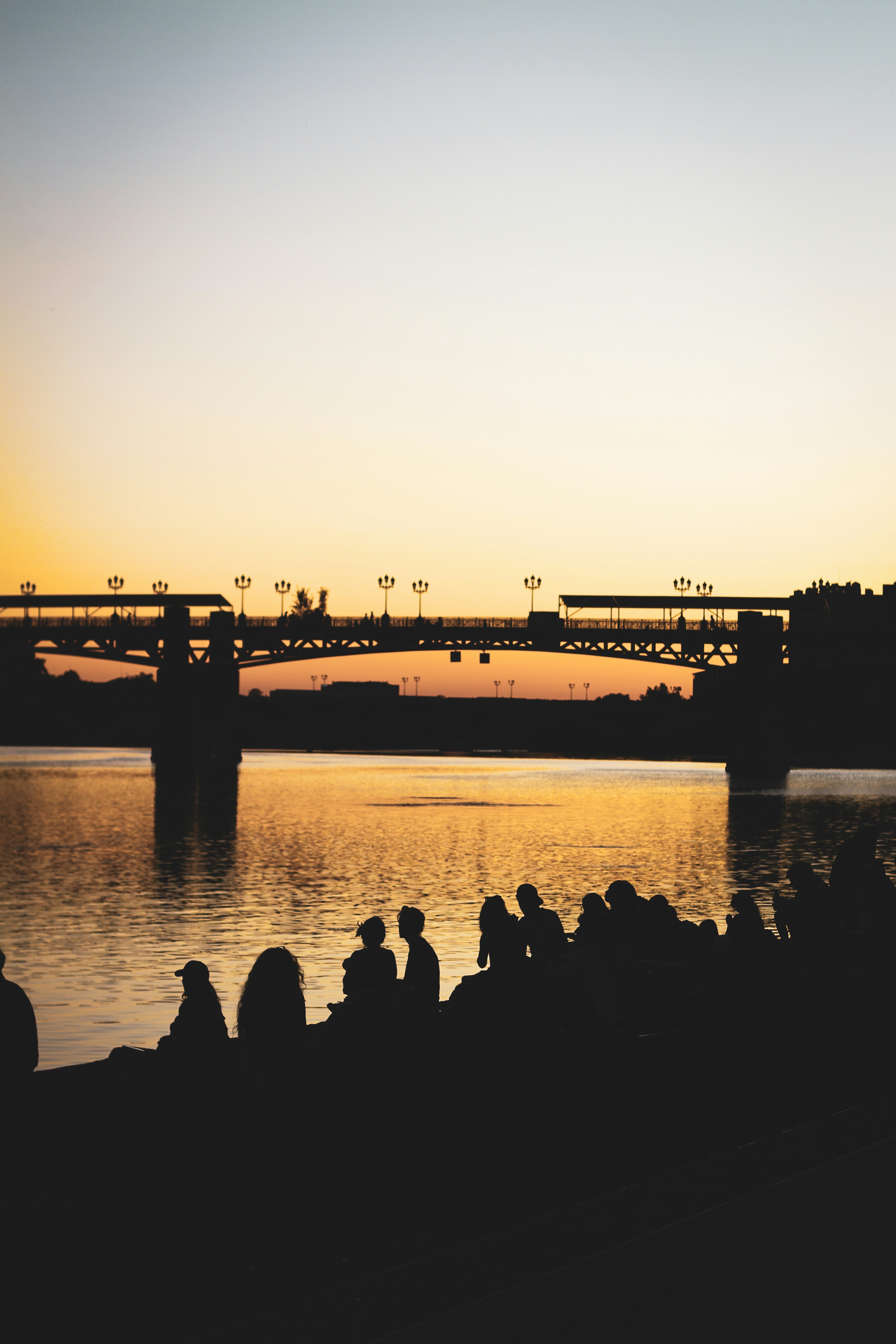 Last Light Over the River: Silhouettes Gather Under a Distant Bridge at Dusk