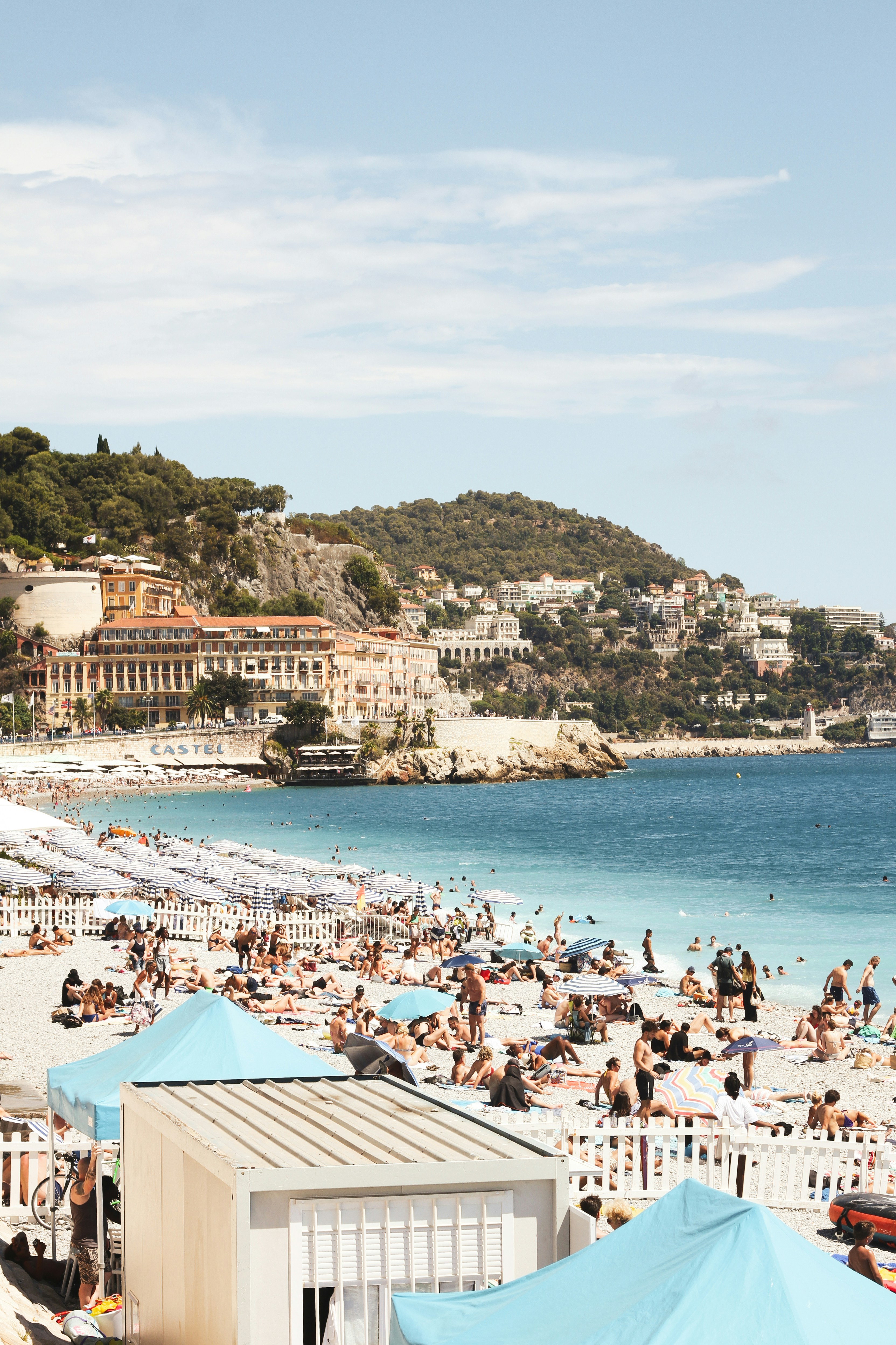 Vibrant beach scene showcasing sunbathers and colorful umbrellas along a picturesque coastline. The backdrop features charming buildings nestled against lush hills.