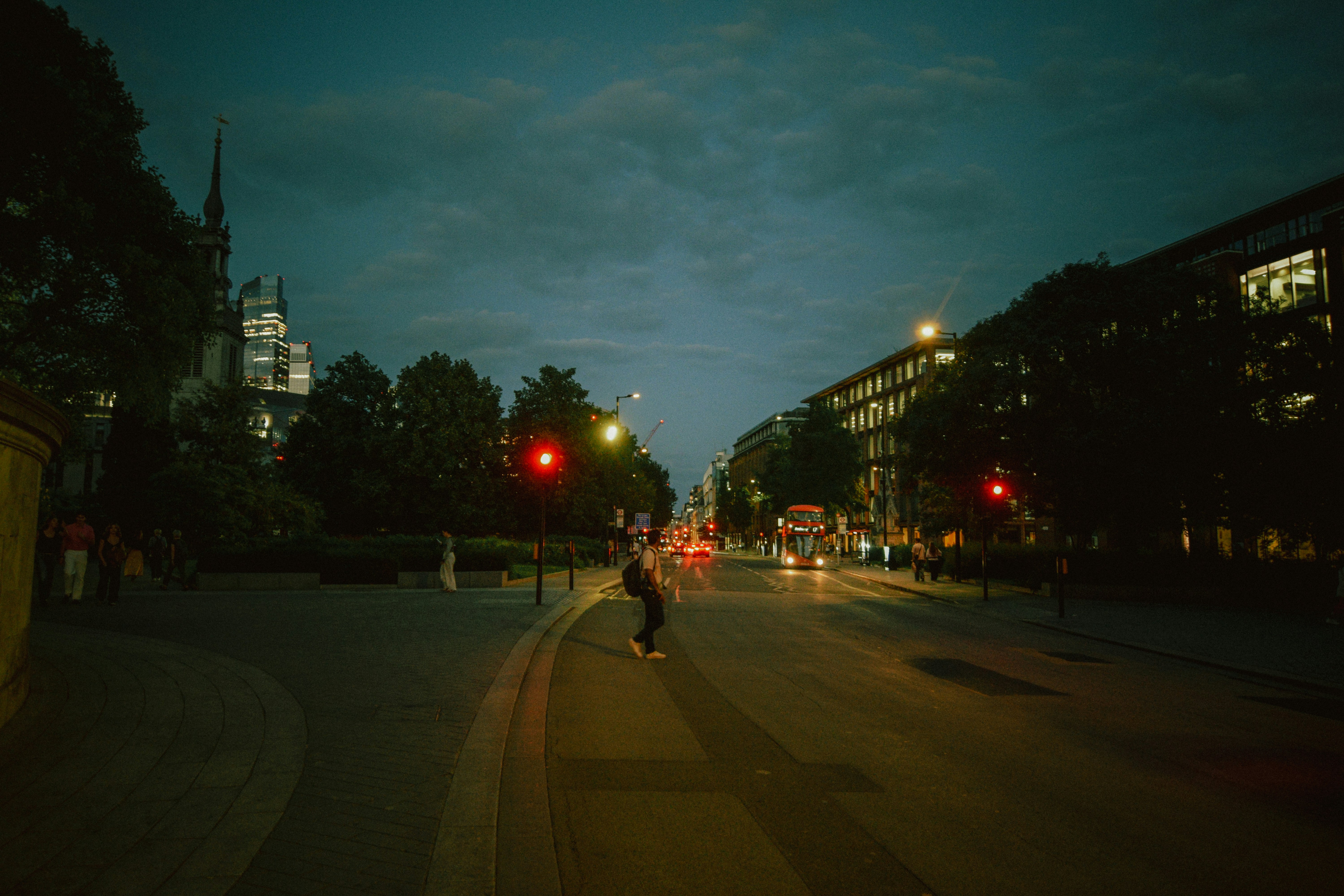 A lone figure walks along a city street illuminated by traffic lights, surrounded by trees and buildings under a twilight sky.