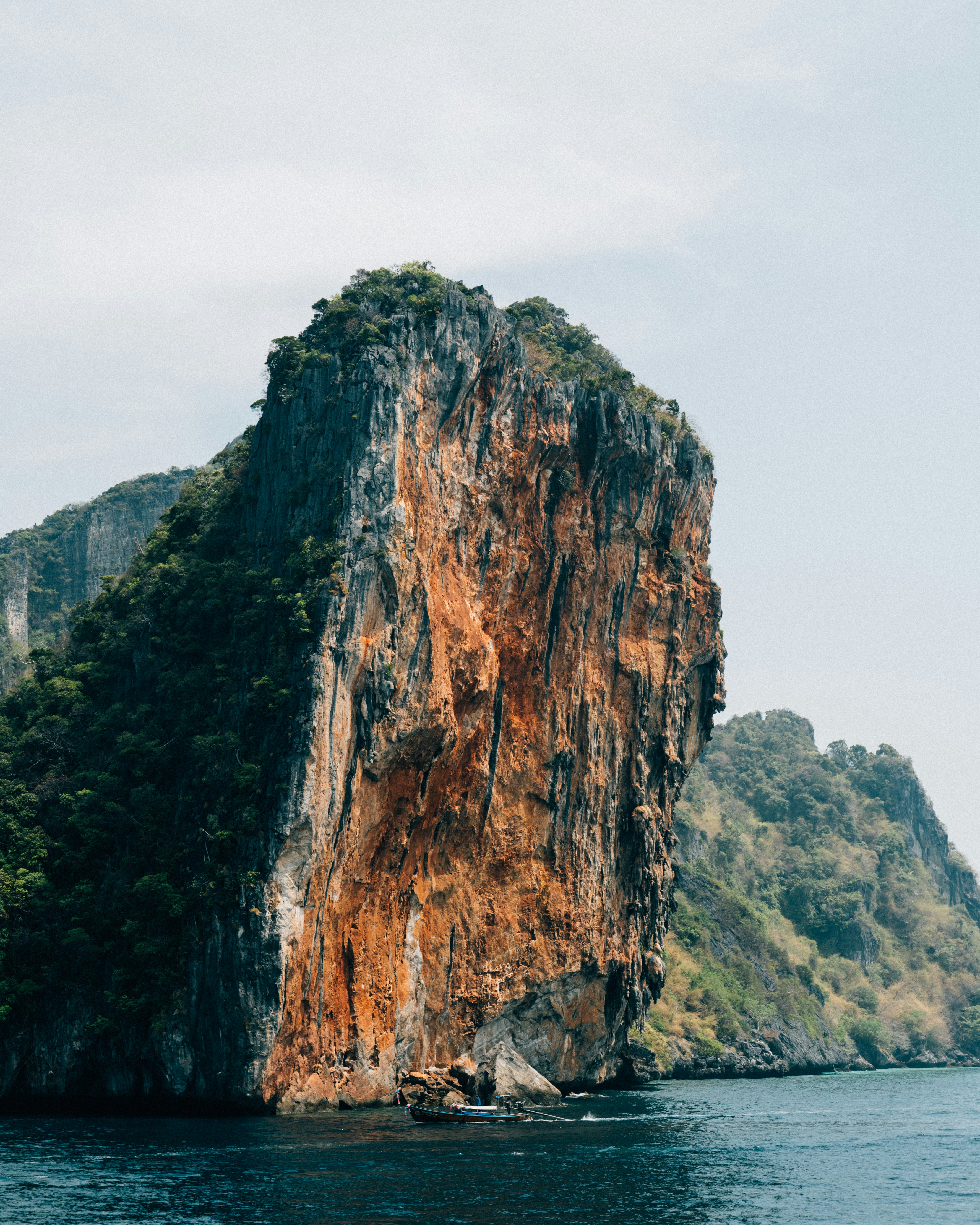 Towering limestone cliff adorned with lush greenery, reflecting the serene waters below. A small boat navigates the calm sea, adding scale to the natural wonder.