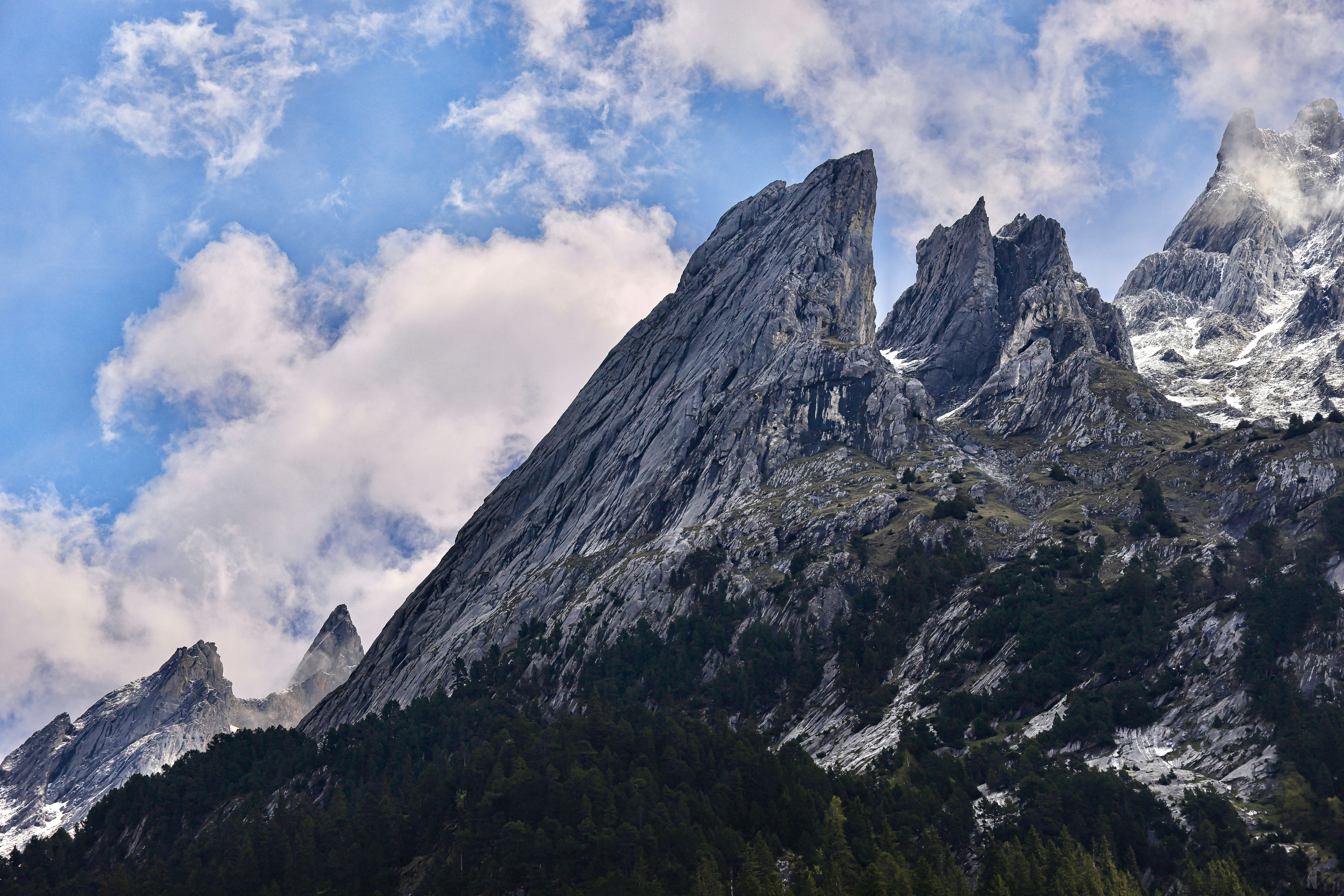 Jagged mountain peaks pierce the sky, surrounded by lush greenery and wispy clouds. A striking display of nature's grandeur.